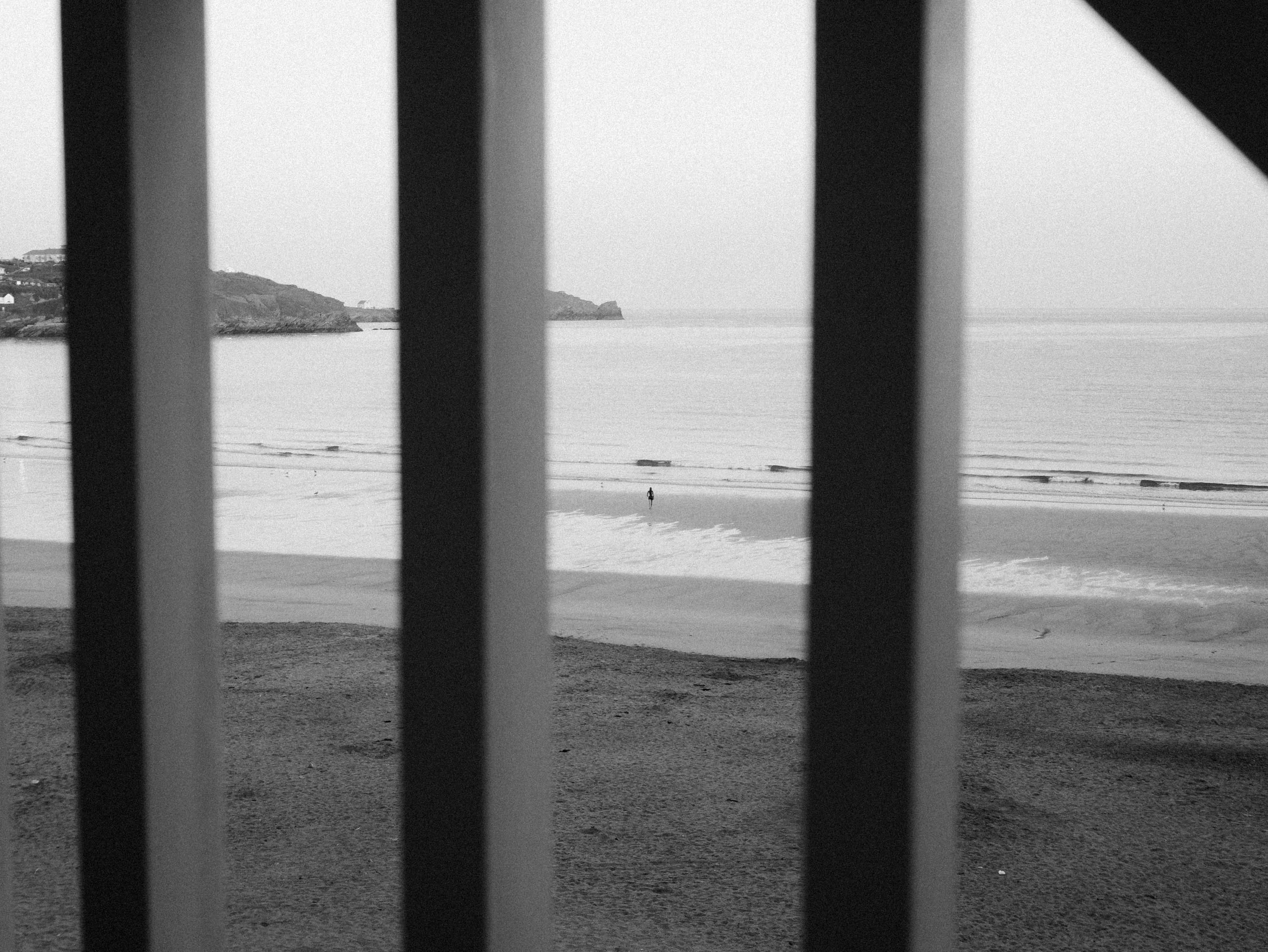 Black-and-white coastal scene seen through vertical bars, with distant shoreline and calm water; a minimal composition emphasizing negative space and rhythm of the bars.