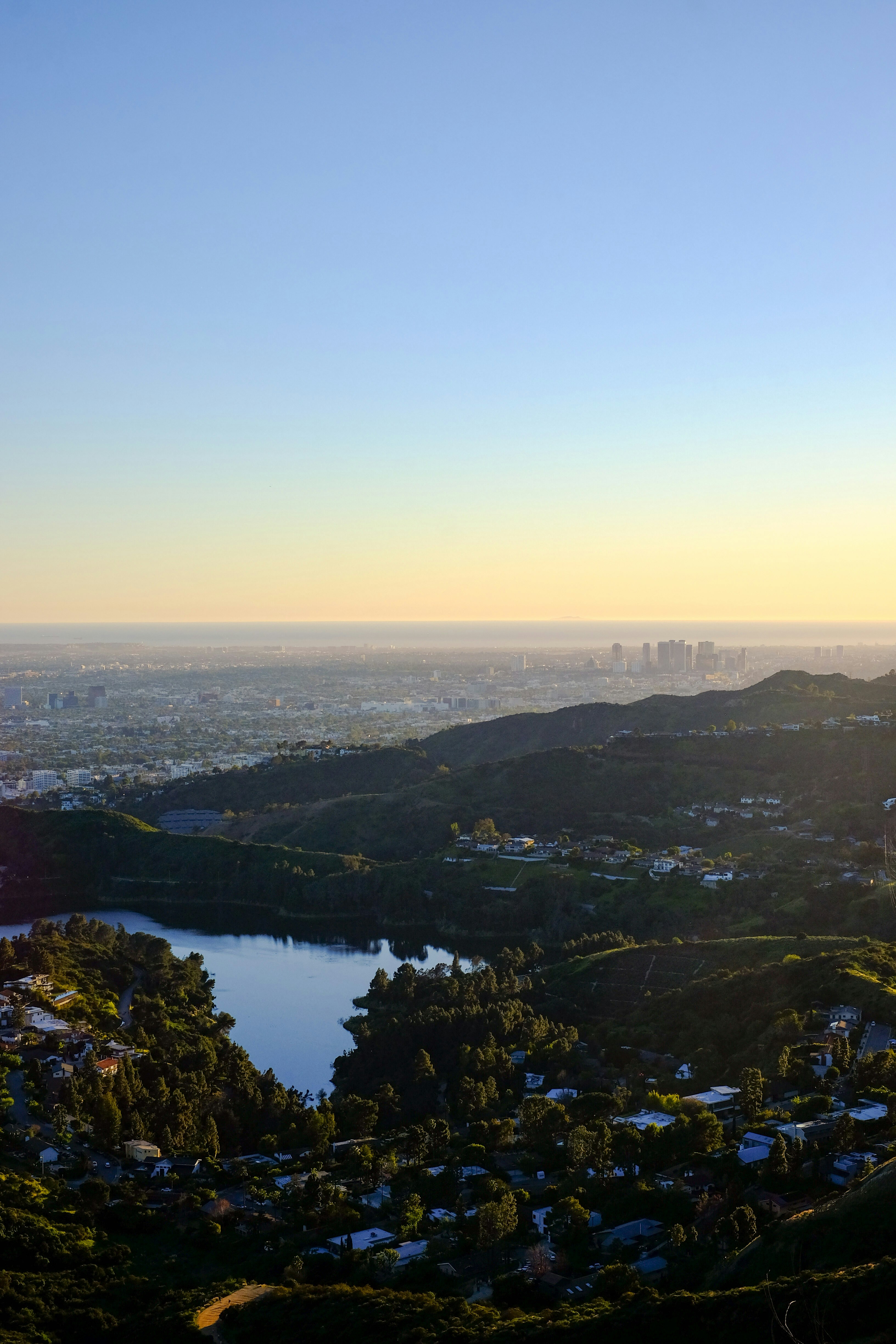 Twilight over a hillside suburb with a distant city skyline and a tranquil lake reflecting the fading light.