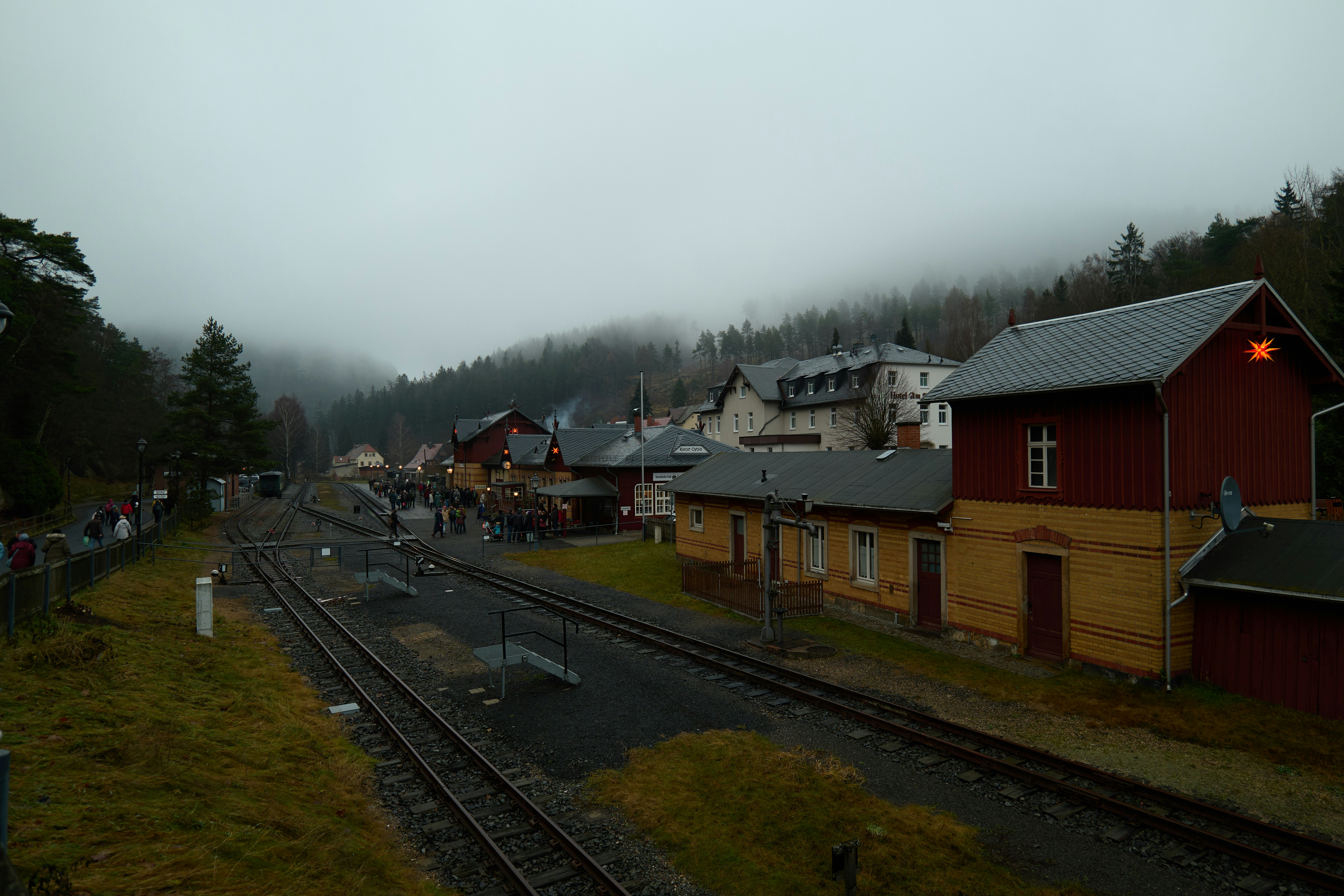 Dim, misty scene of a small railway yard with colorful wooden buildings along the tracks and distant fog-shrouded hills.