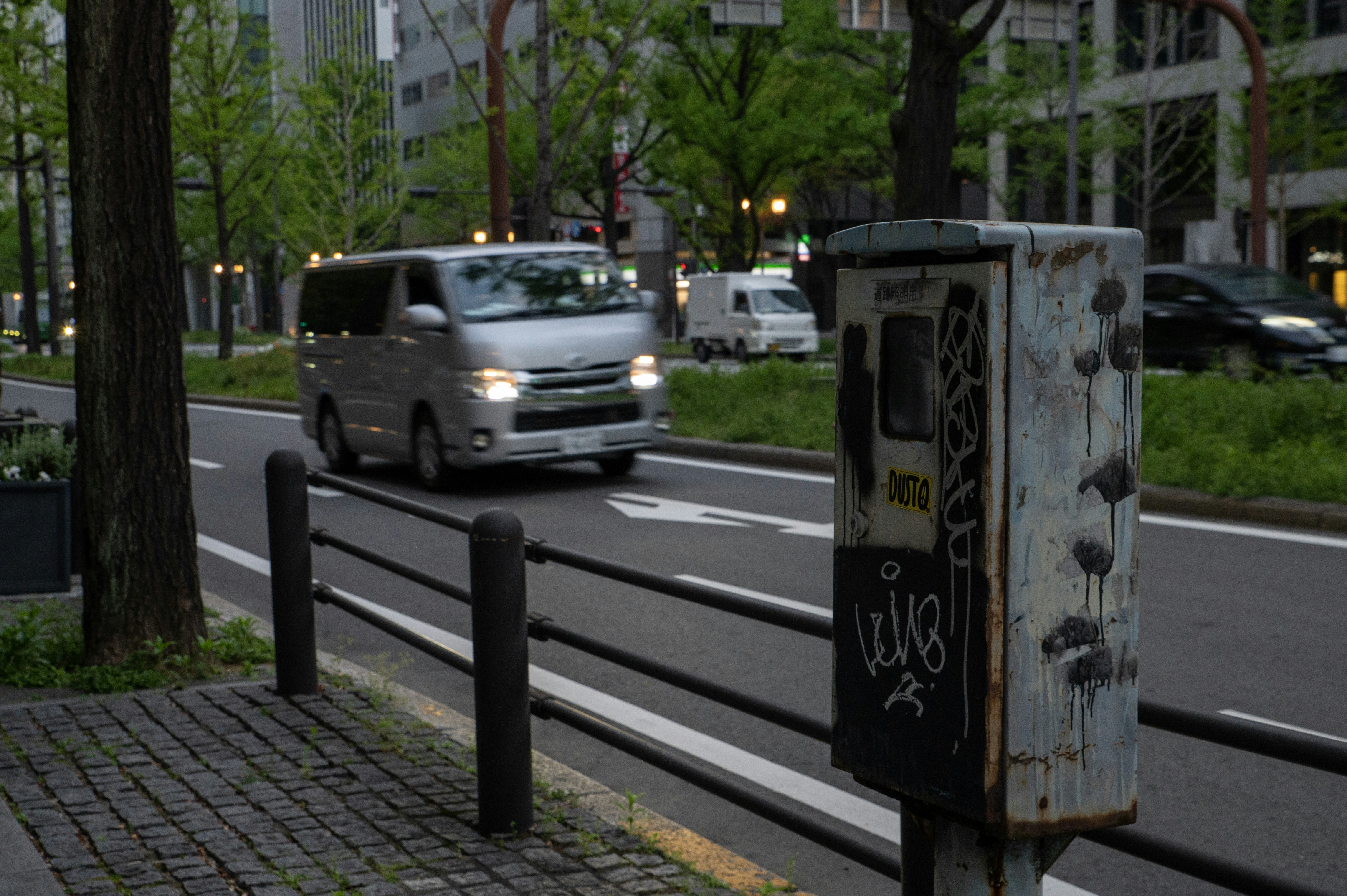 Streetscale: A rusted box on a quiet urban curb watches over passing traffic