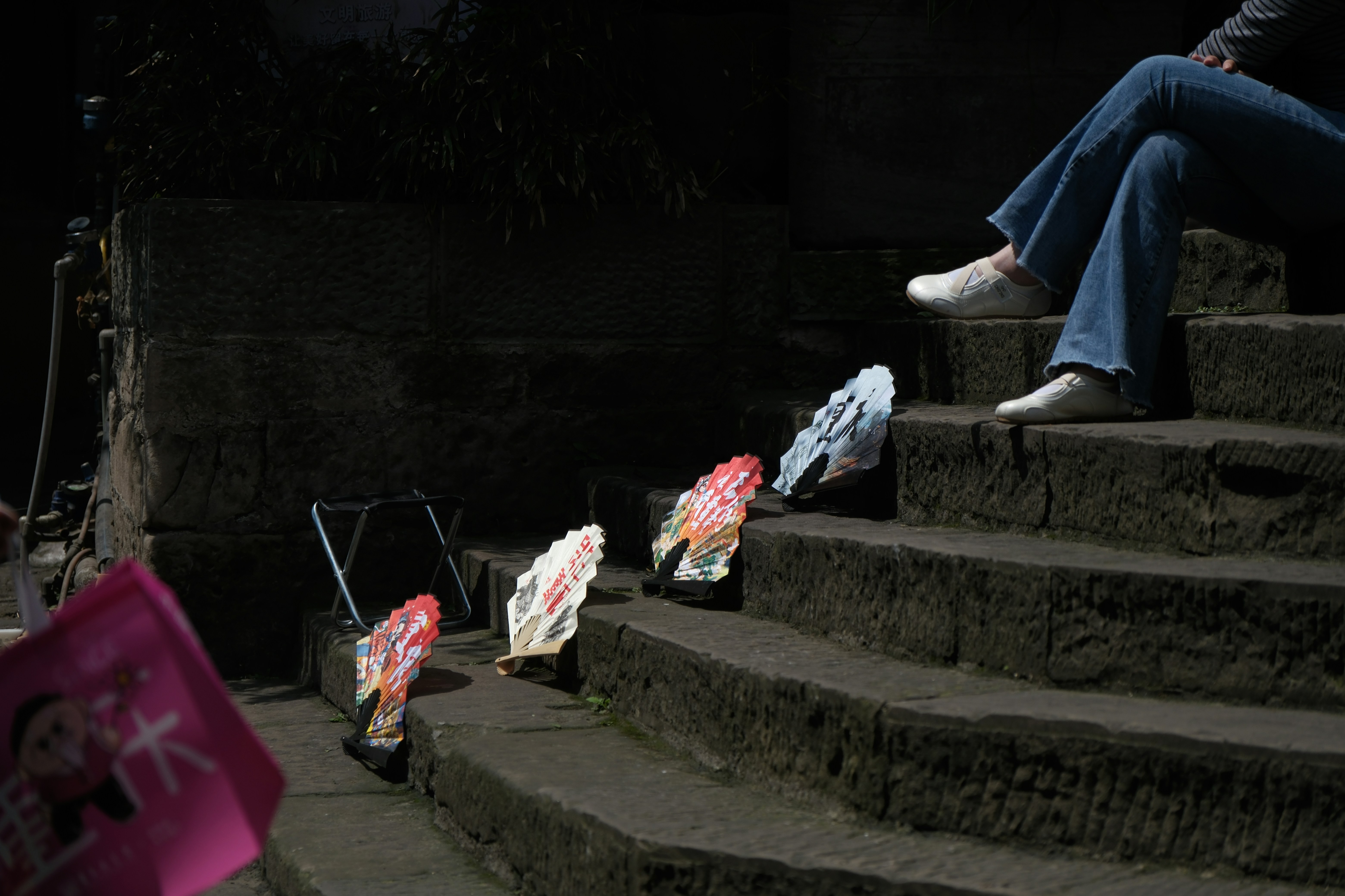 Colorful paper cutouts line the stone steps as a person sits at the top, their legs in blue jeans and white sneakers visible. The scene emphasizes texture of the steps and subtle ambient light.
