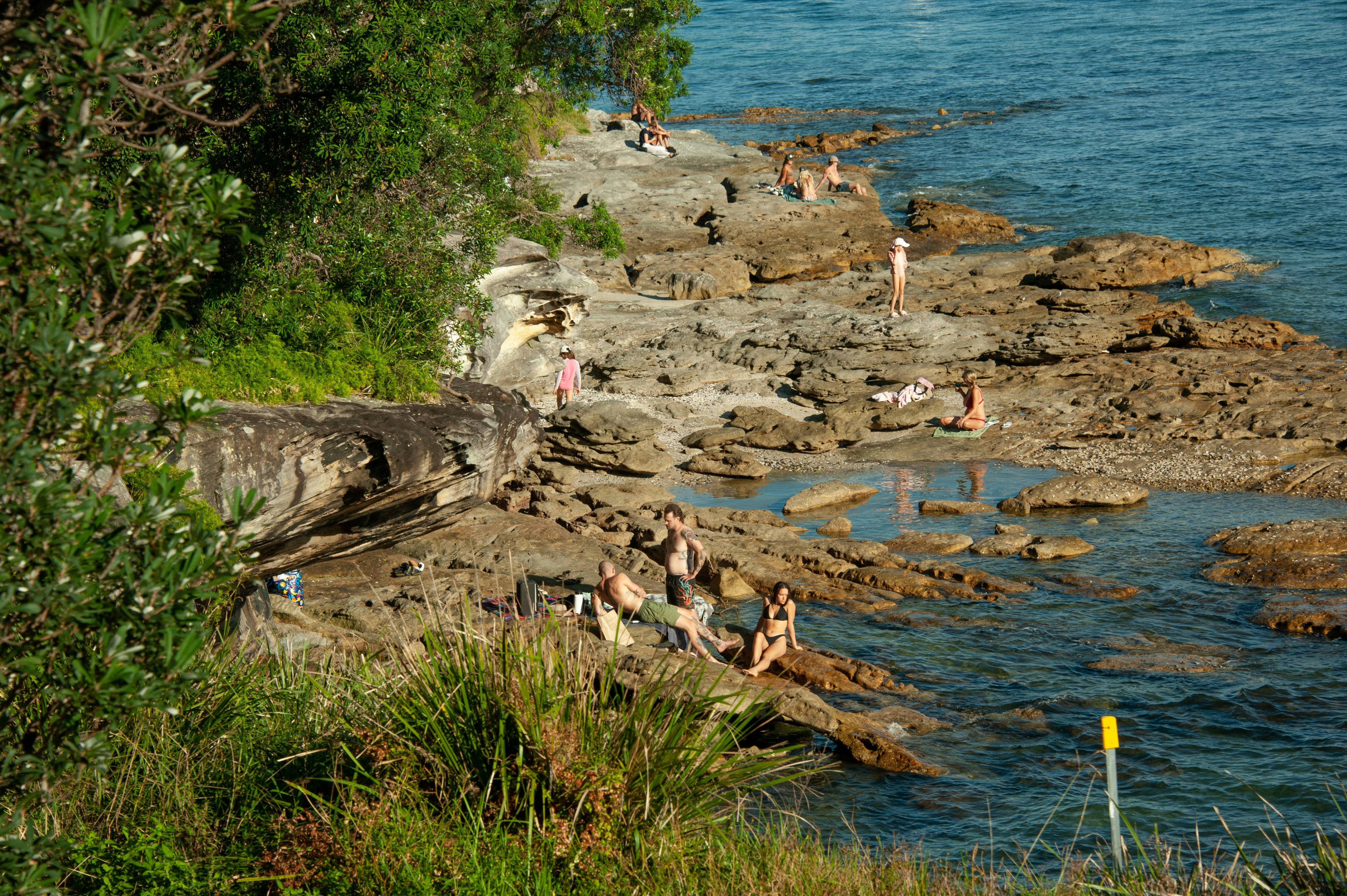 Sunlit Cove: Beachgoers on Tumbled Rocks by a Quiet Sea