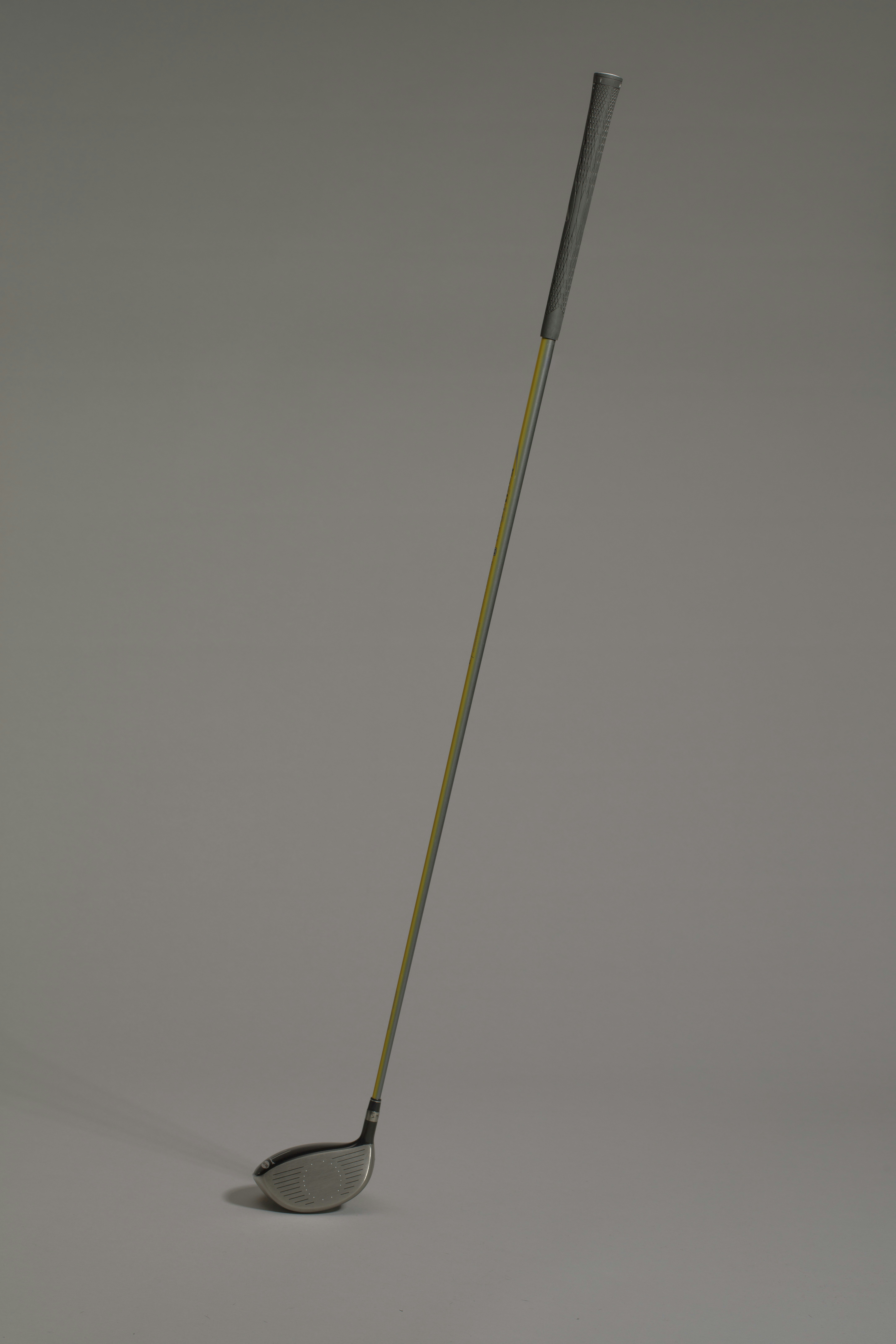 A single golf club rests on a neutral backdrop, showcasing a long shaft and a rounded clubhead with a subtle shadow to the left. The image highlights the equipment’s form and texture without surrounding context.