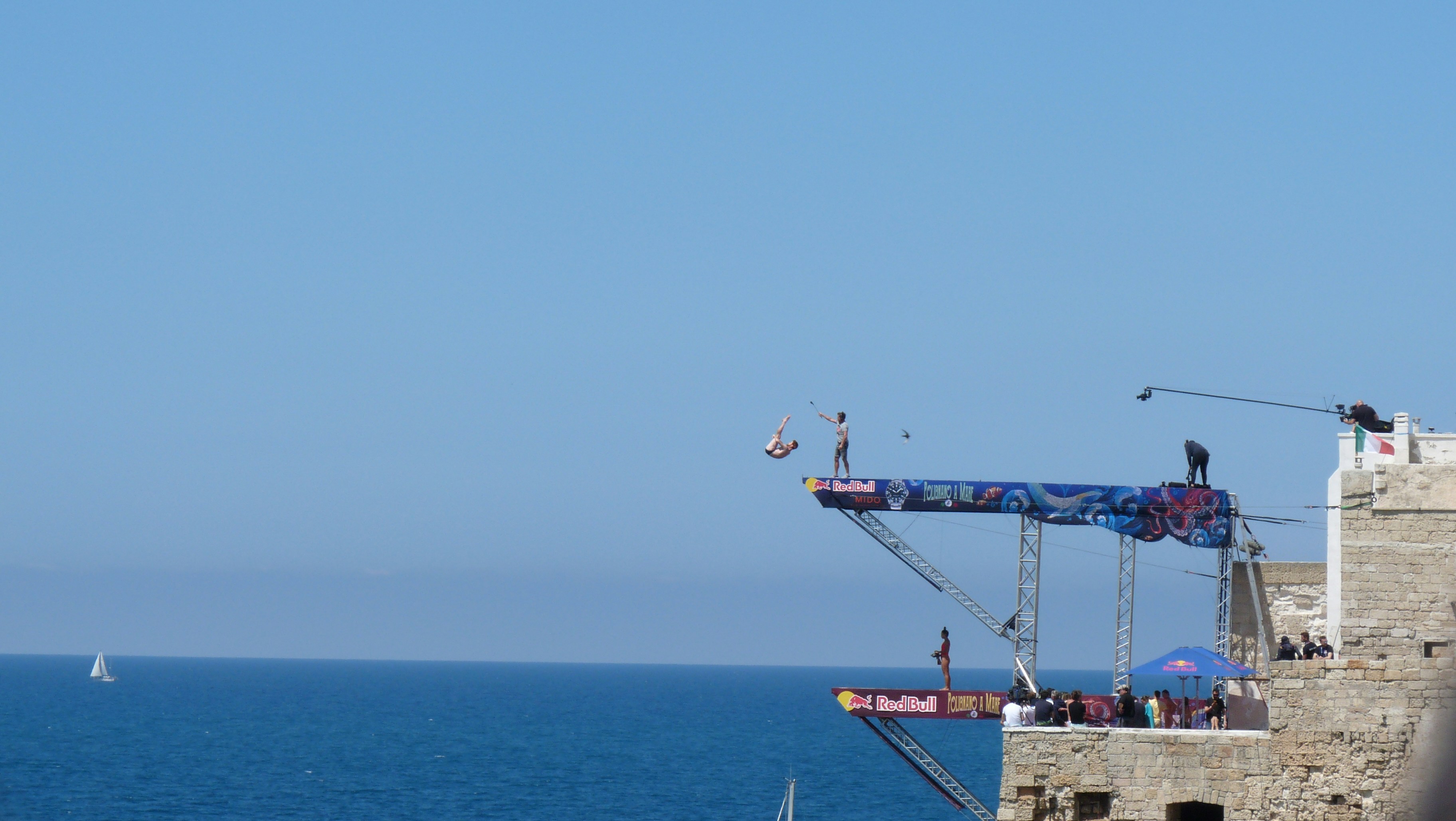 People leap from a coastal diving platform into calm blue water under a clear sky, with a distant sailboat on the horizon.