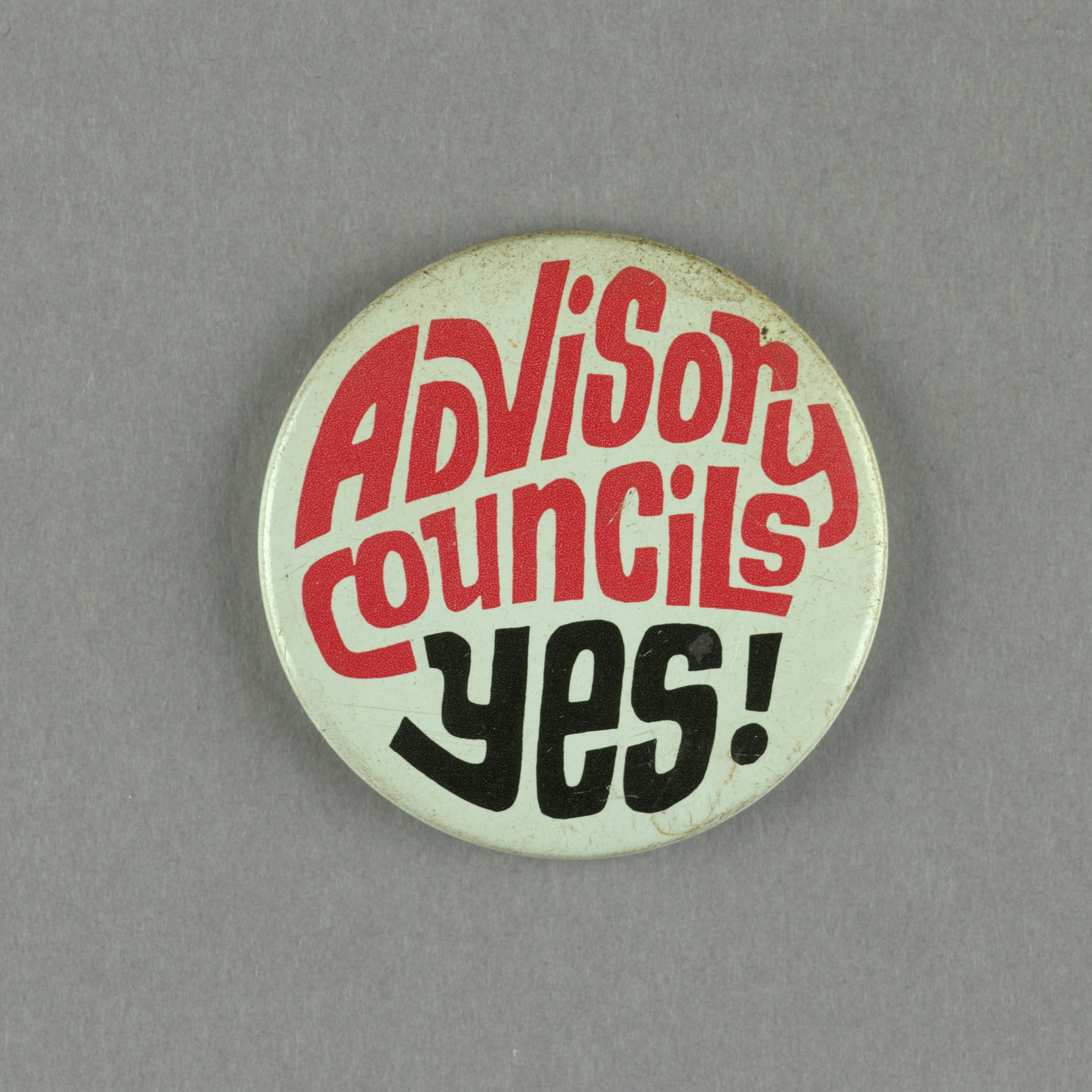Round badge with red/black 'Advisory Councils' typography on pale background; a political pin resting on plain gray surface.