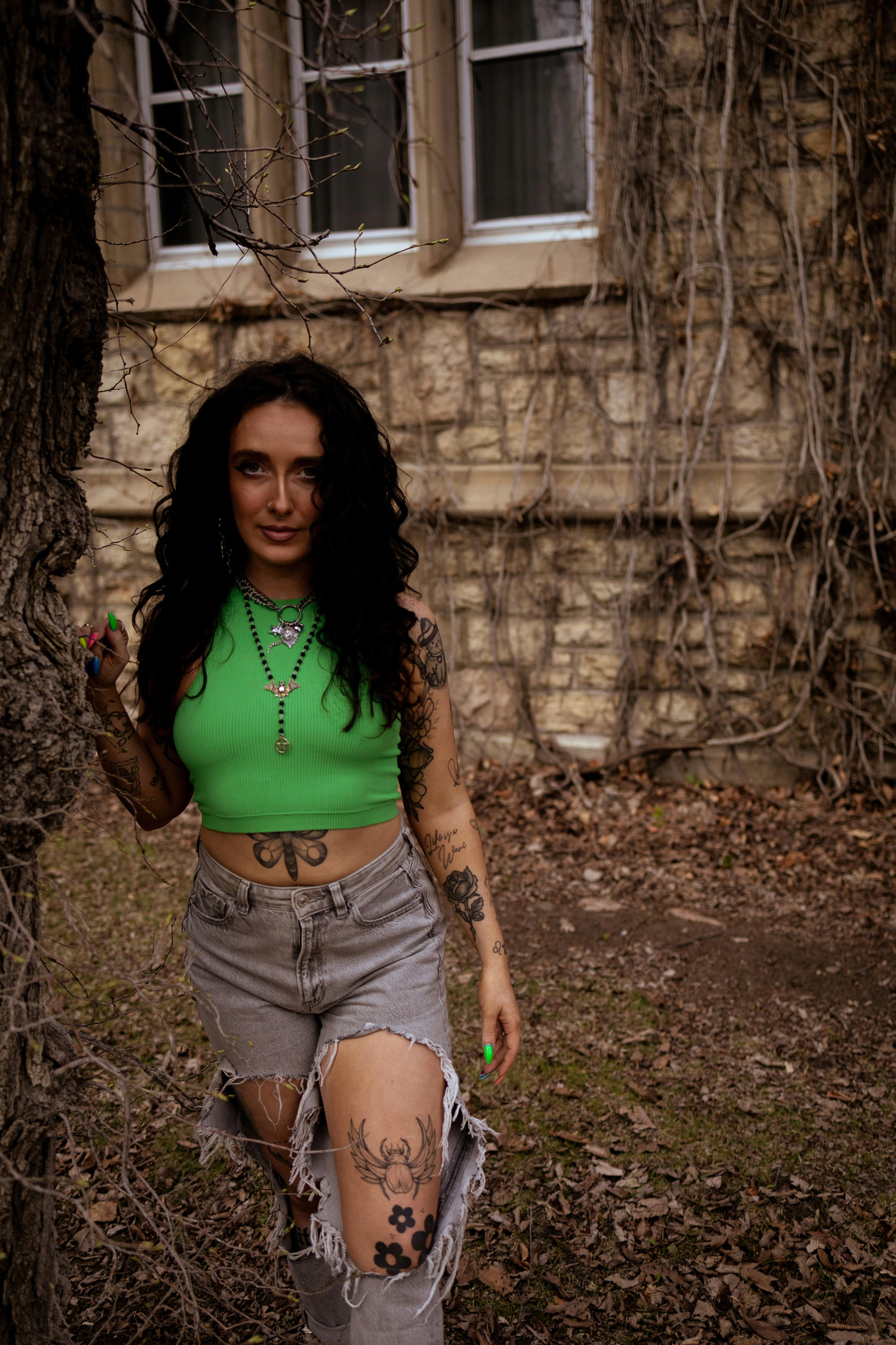 Woman in a bright green crop top and ripped denim walks among ivy-covered stone walls; tattoos visible on arms and torso, natural outdoor setting. (Logo-free photograph).