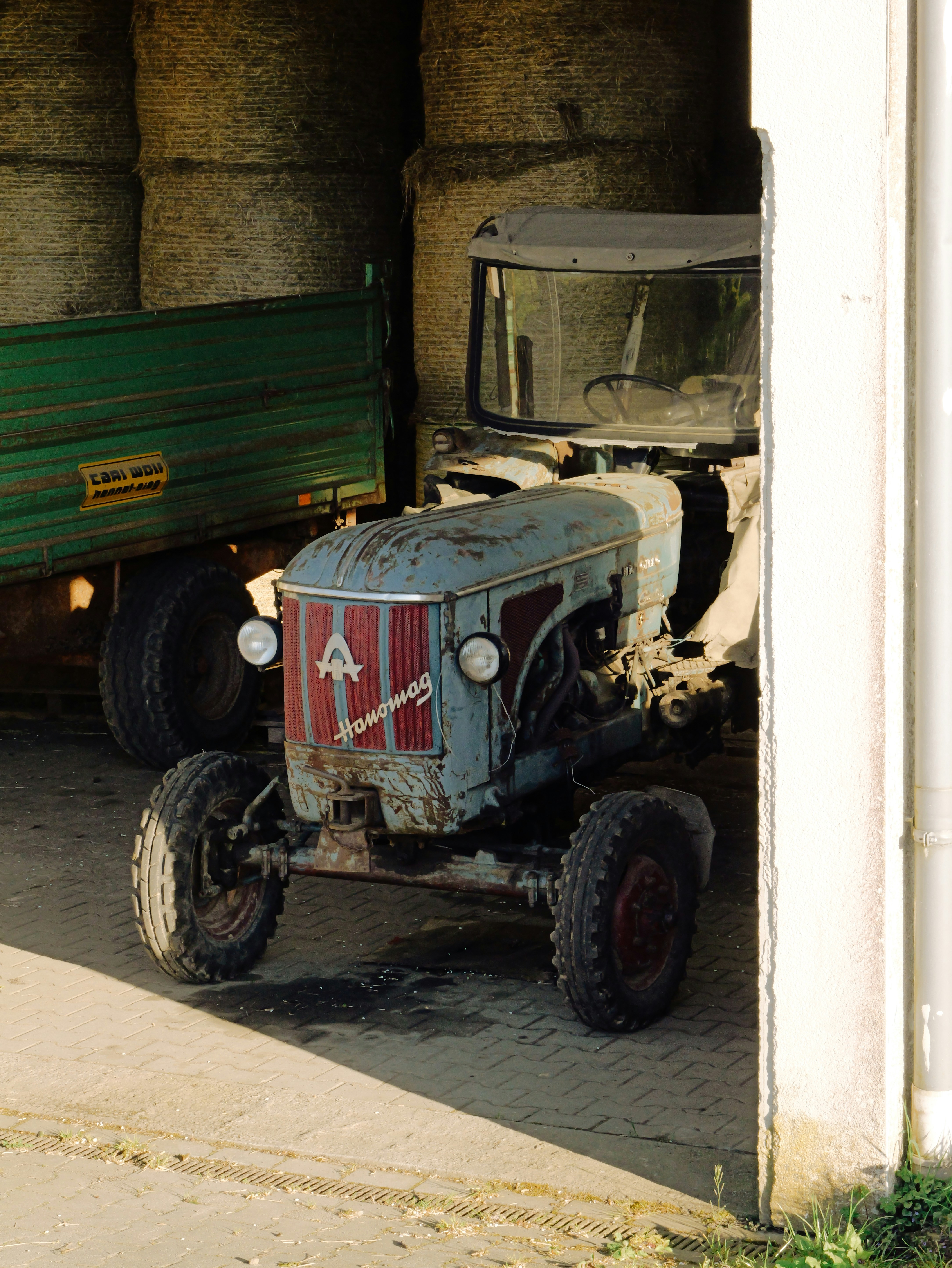 Dusty blue Ferguson tractor tucked inside a barn with a green trailer