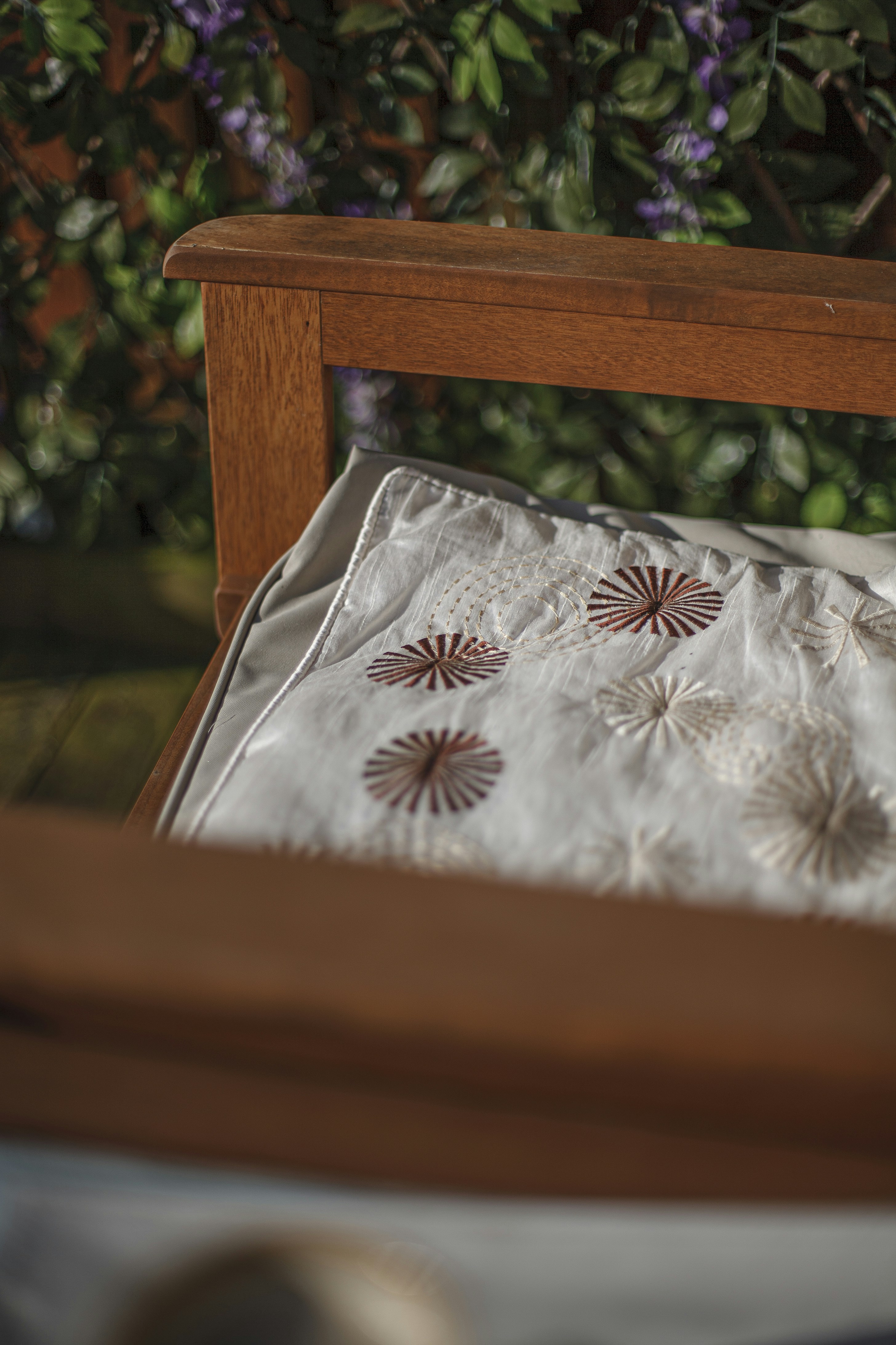 Wooden chair with a patterned cushion sits in soft outdoor light, plants in the background adding depth and color.