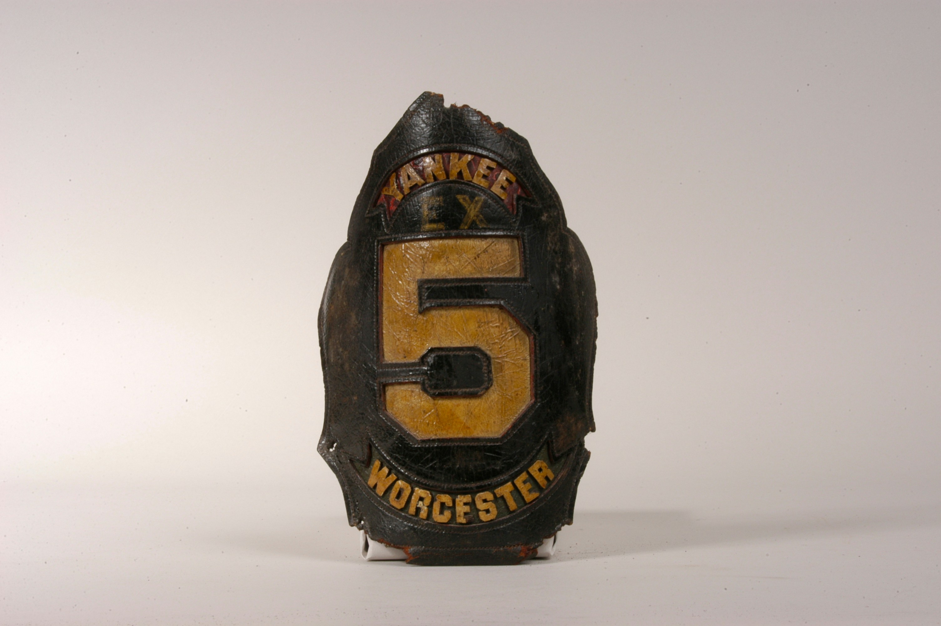 A worn vintage badge with a bold yellow number 5 sits on a clean pale surface, evoking history and character. This is a small, glossy emblem, captured in a simple studio setup.