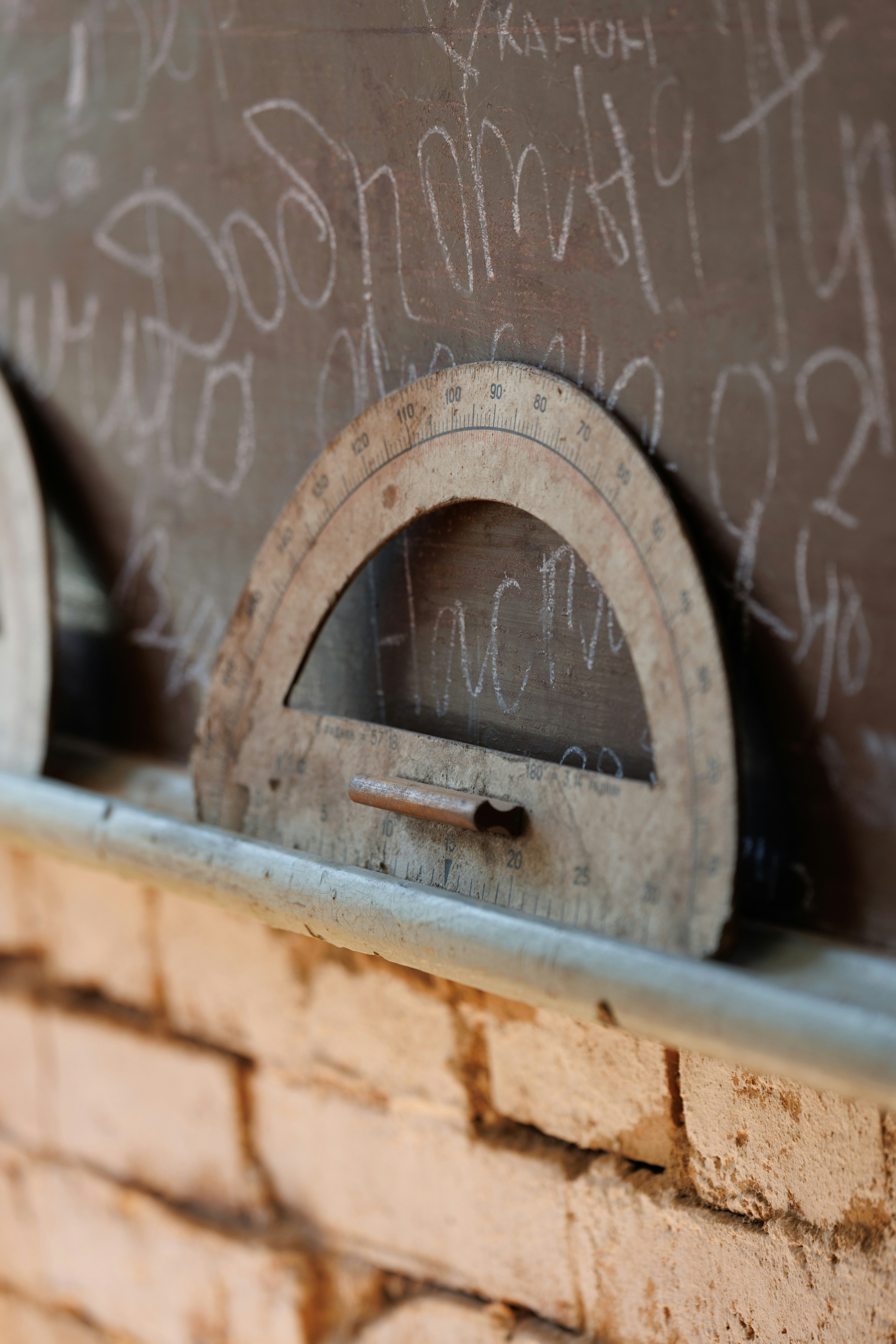 Rusty semicircular protractor on a chalkboard, with chalk markings and a brick windowsill in the foreground.