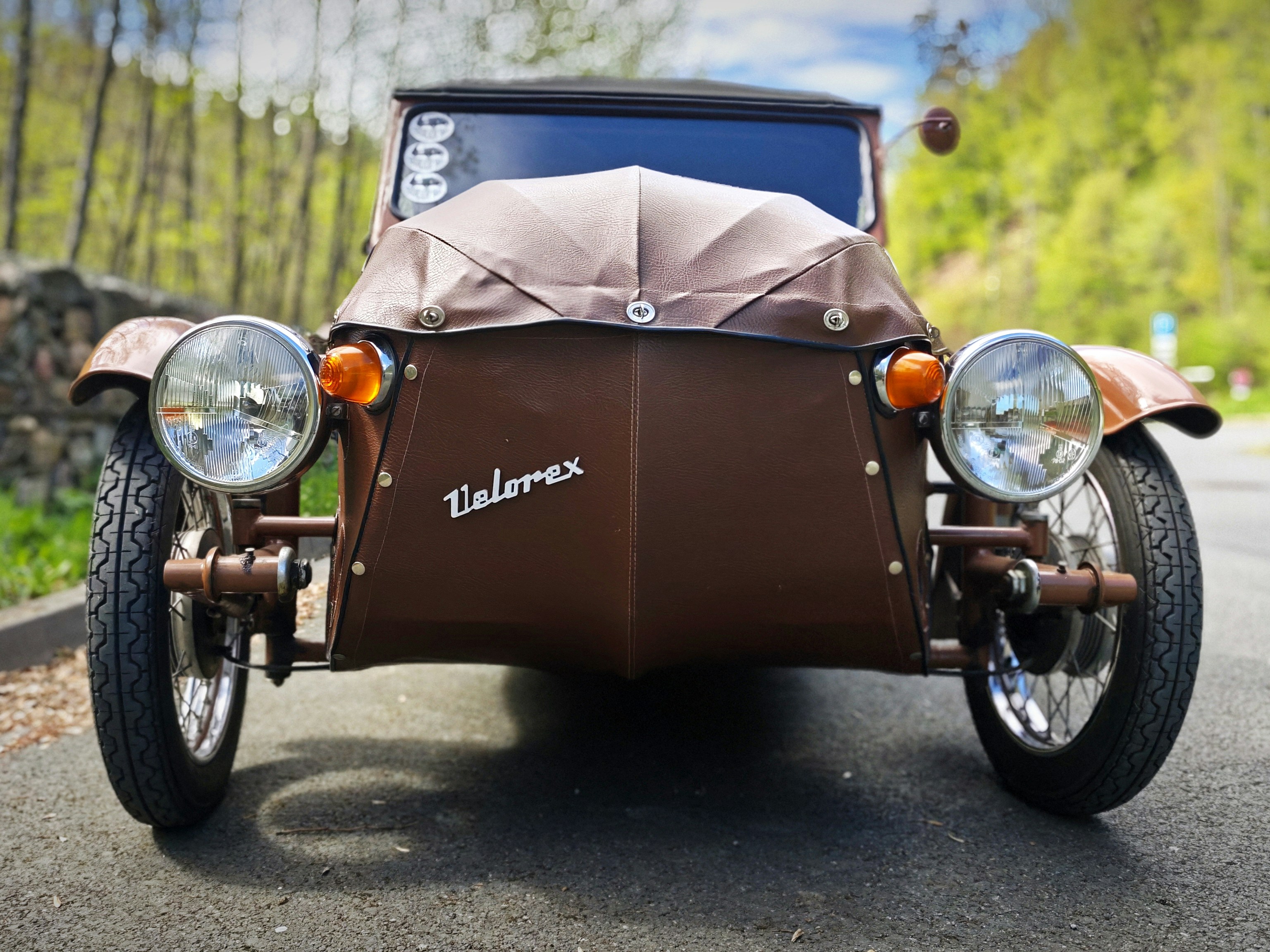 Brown vintage car with a folded canopy, round headlights, and exposed wheels parked on a paved road amid trees.