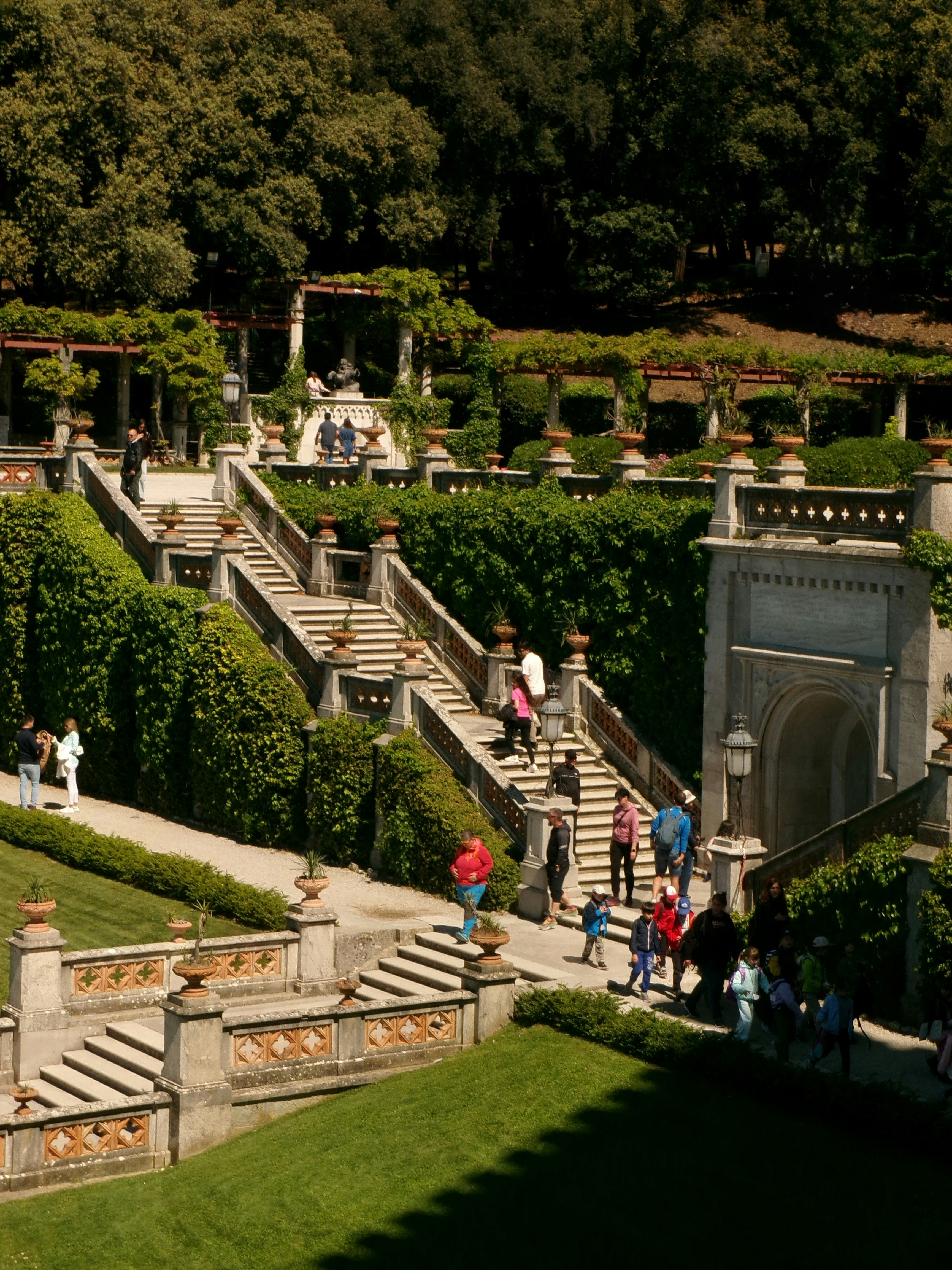 Terraced stone stairs wind through a manicured garden with hedges and visitors exploring the pathways; architectural arches and carved balustrades frame the scene.