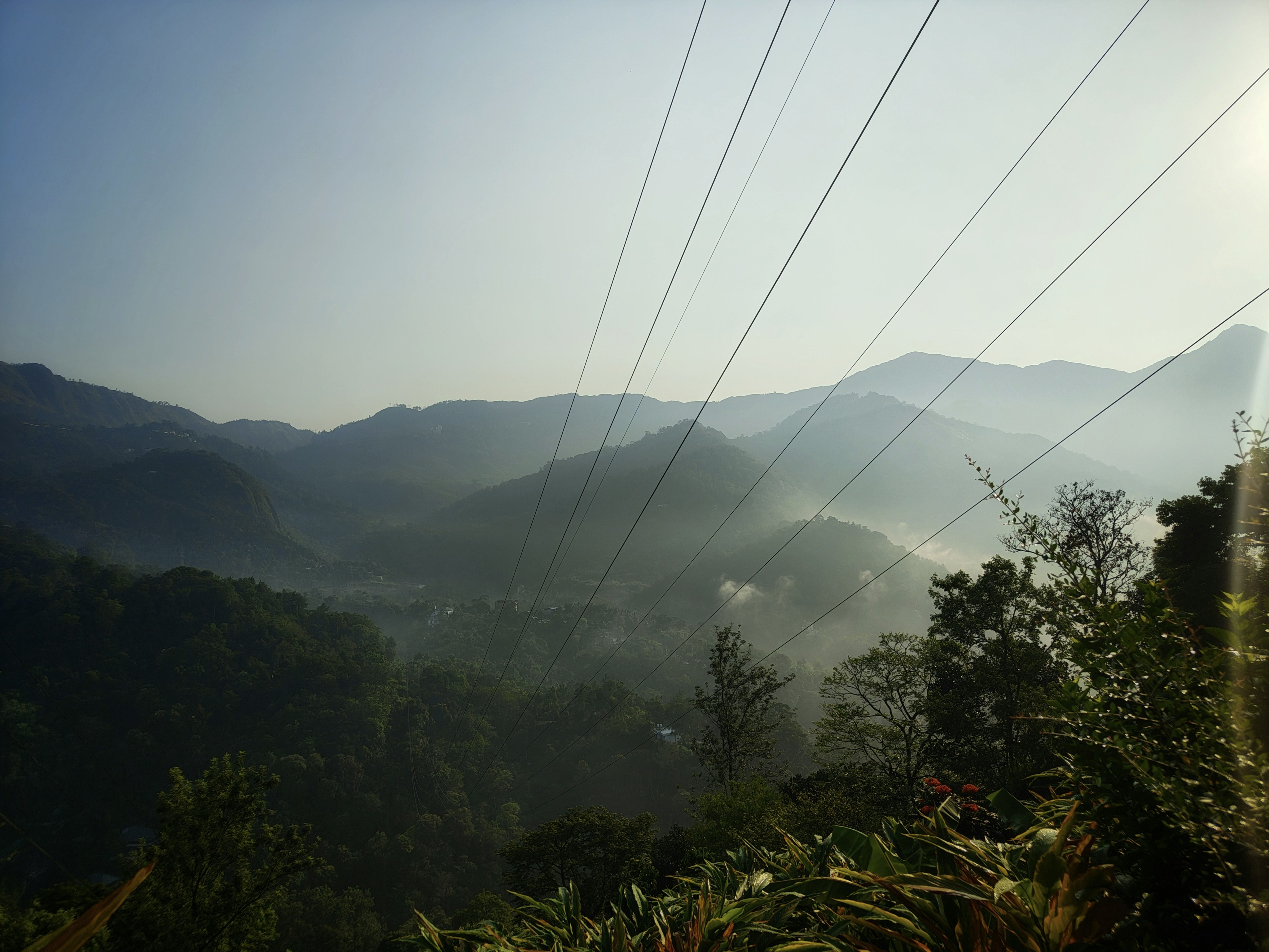 Hazy valley hills fade into the distance with power lines crossing the sky, vegetation in the foreground and soft sunlight below the ridge.