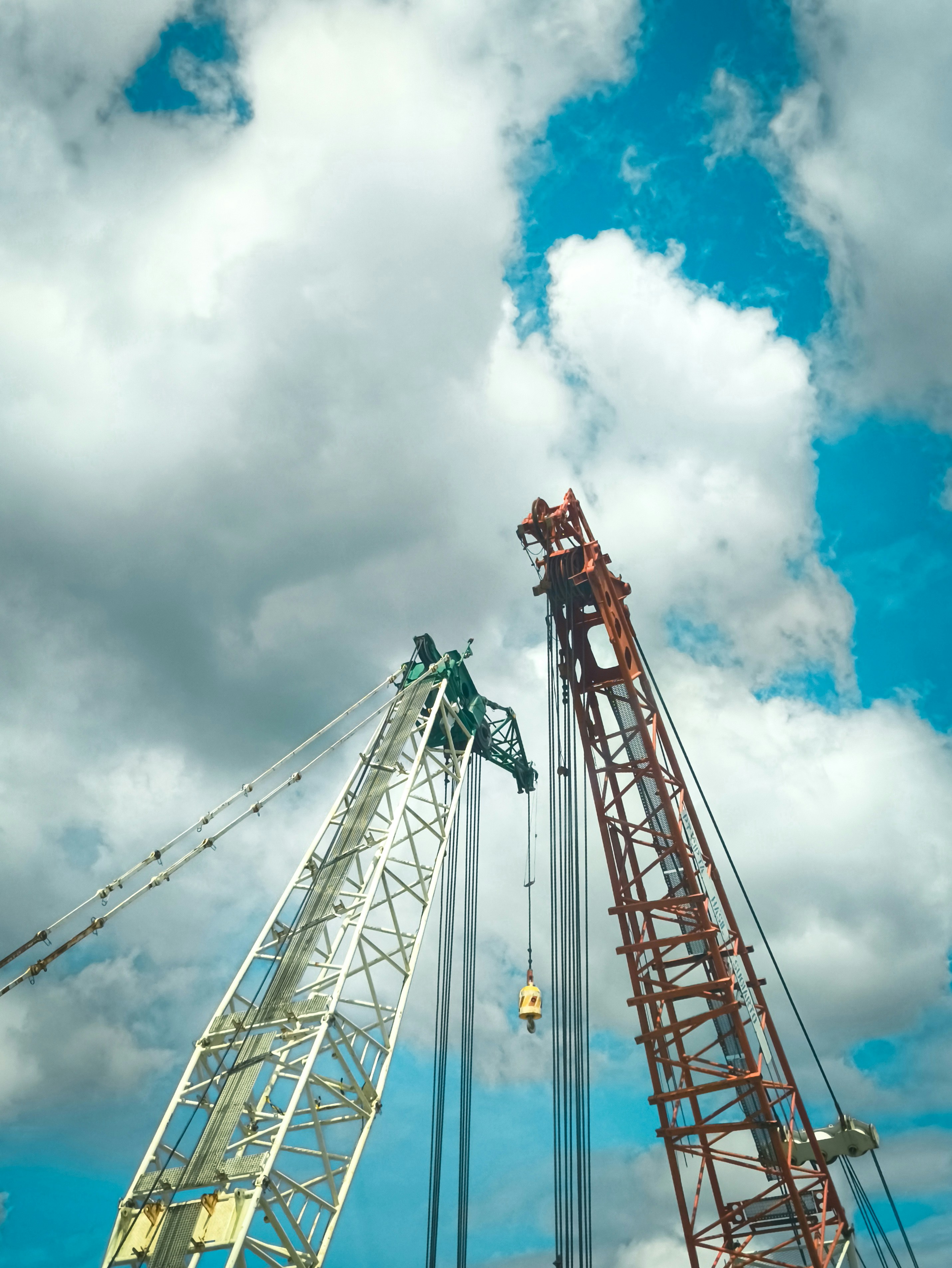 Two construction cranes (white-green and red) tower against a cloudy blue sky. This composition highlights the sturdy steel lattice structures and the industrial aesthetic against nature.