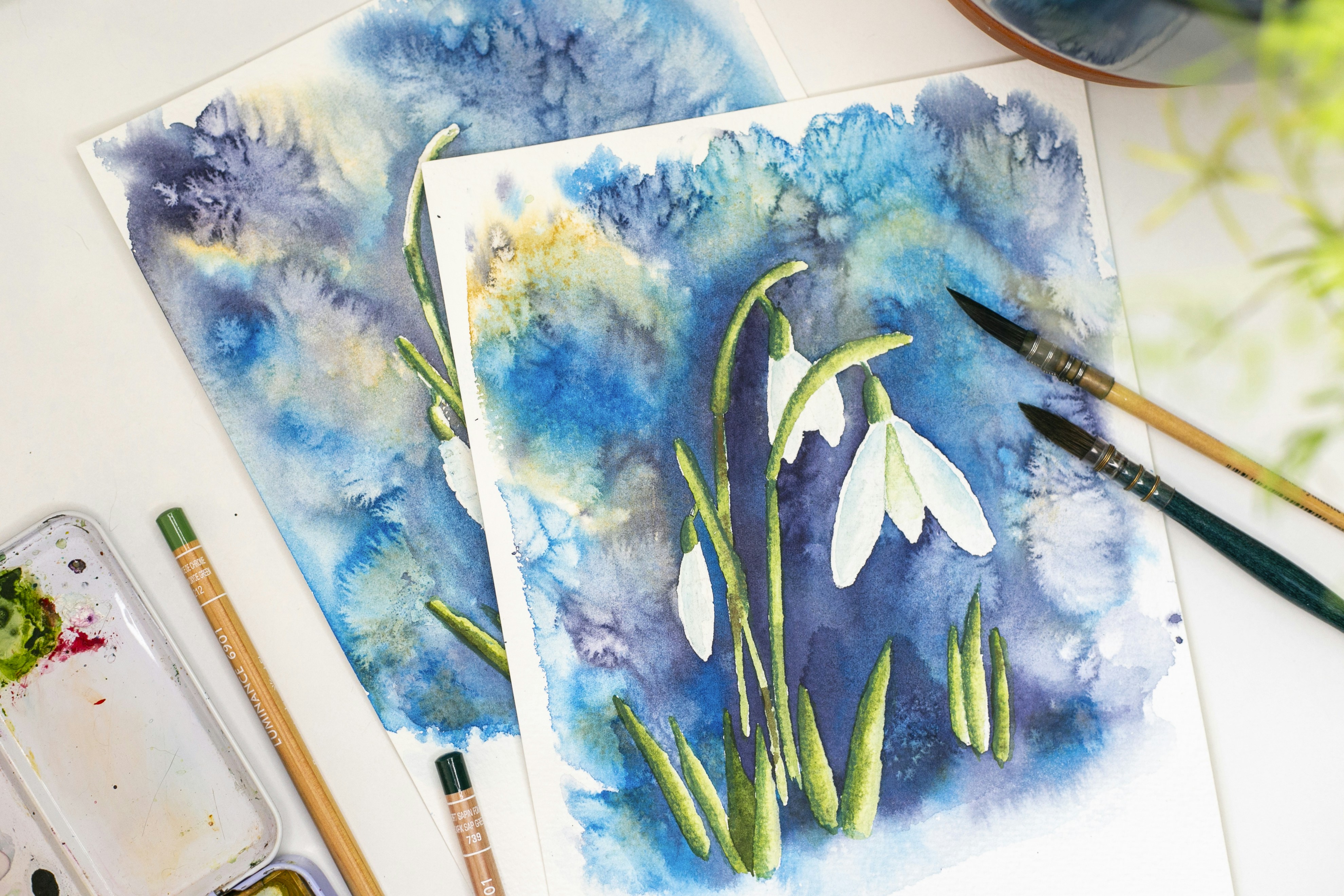 Blue watercolor flowers bend toward sunlight on a white studio table, with paint palettes and brushes nearby; a soft, layered wash creates depth and gentle color transitions. This is a watercolor illustration on paper.
