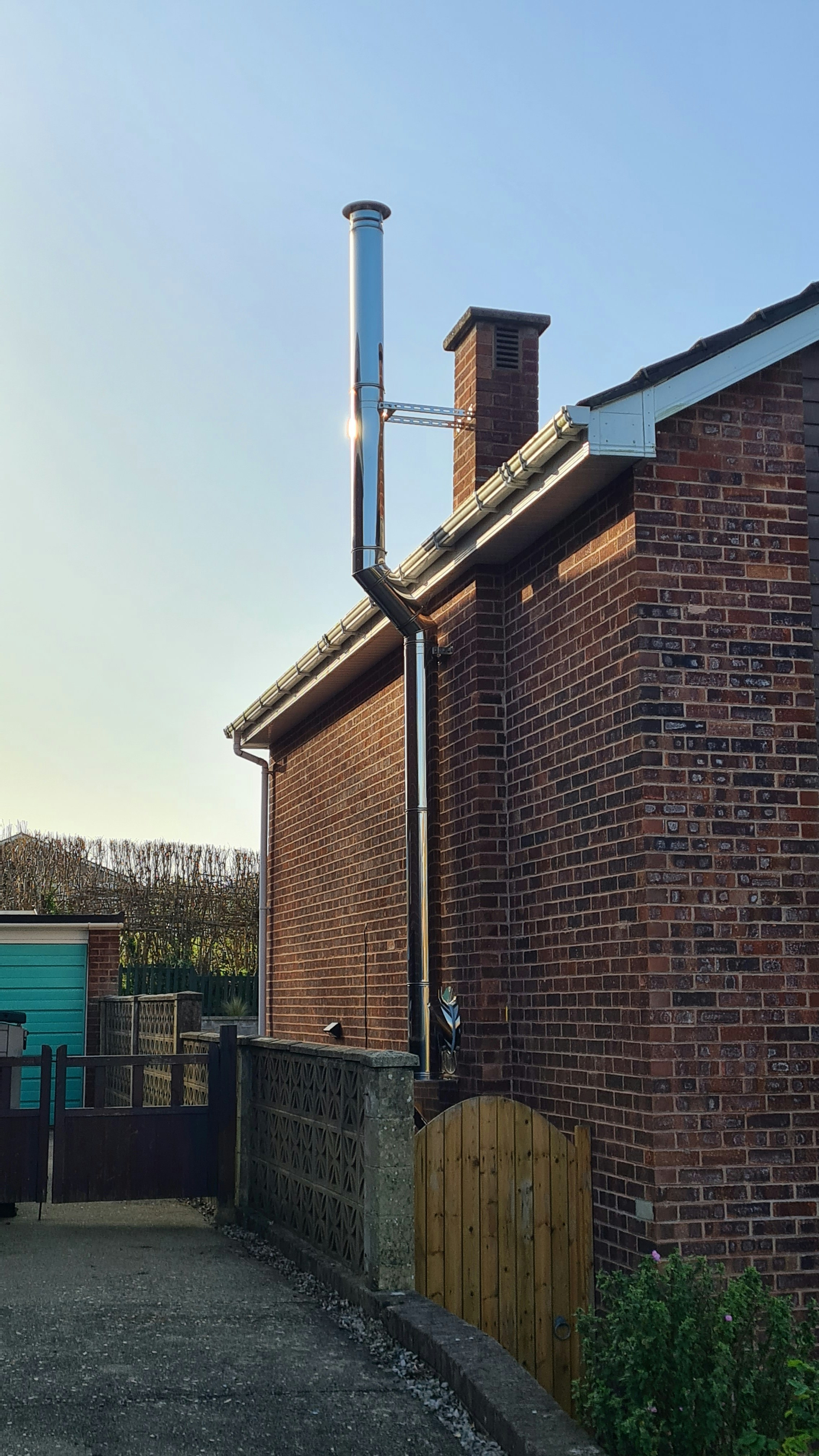 Brick residence with a tall metallic chimney along the side, under a clear blue sky; a gate and fence line the narrow alley beside the building.