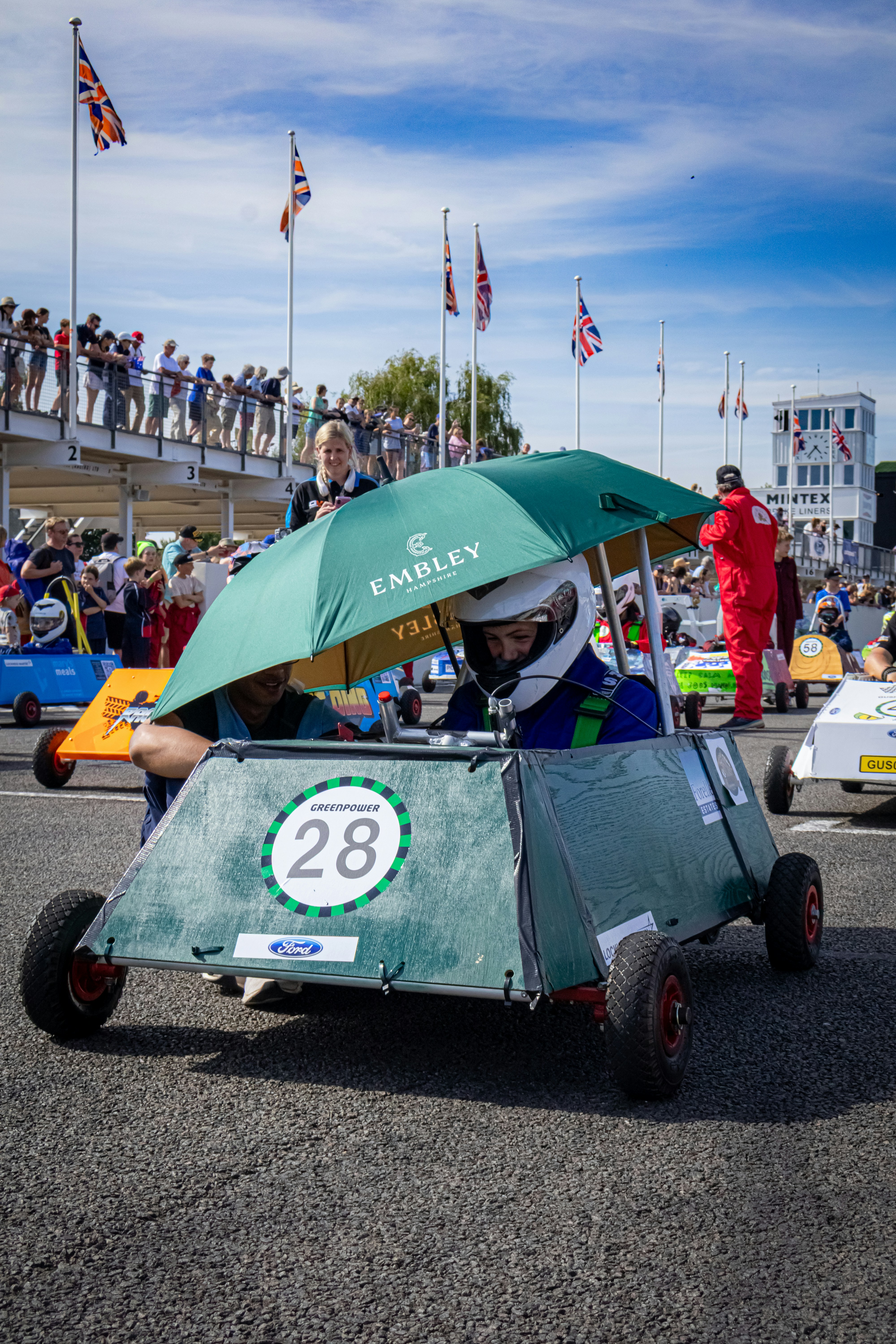 A young driver prepares in the pit lane at a Greenpower electric racing event — helmeted and ready, sheltered under an umbrella as the crowd watches from the bridge above.