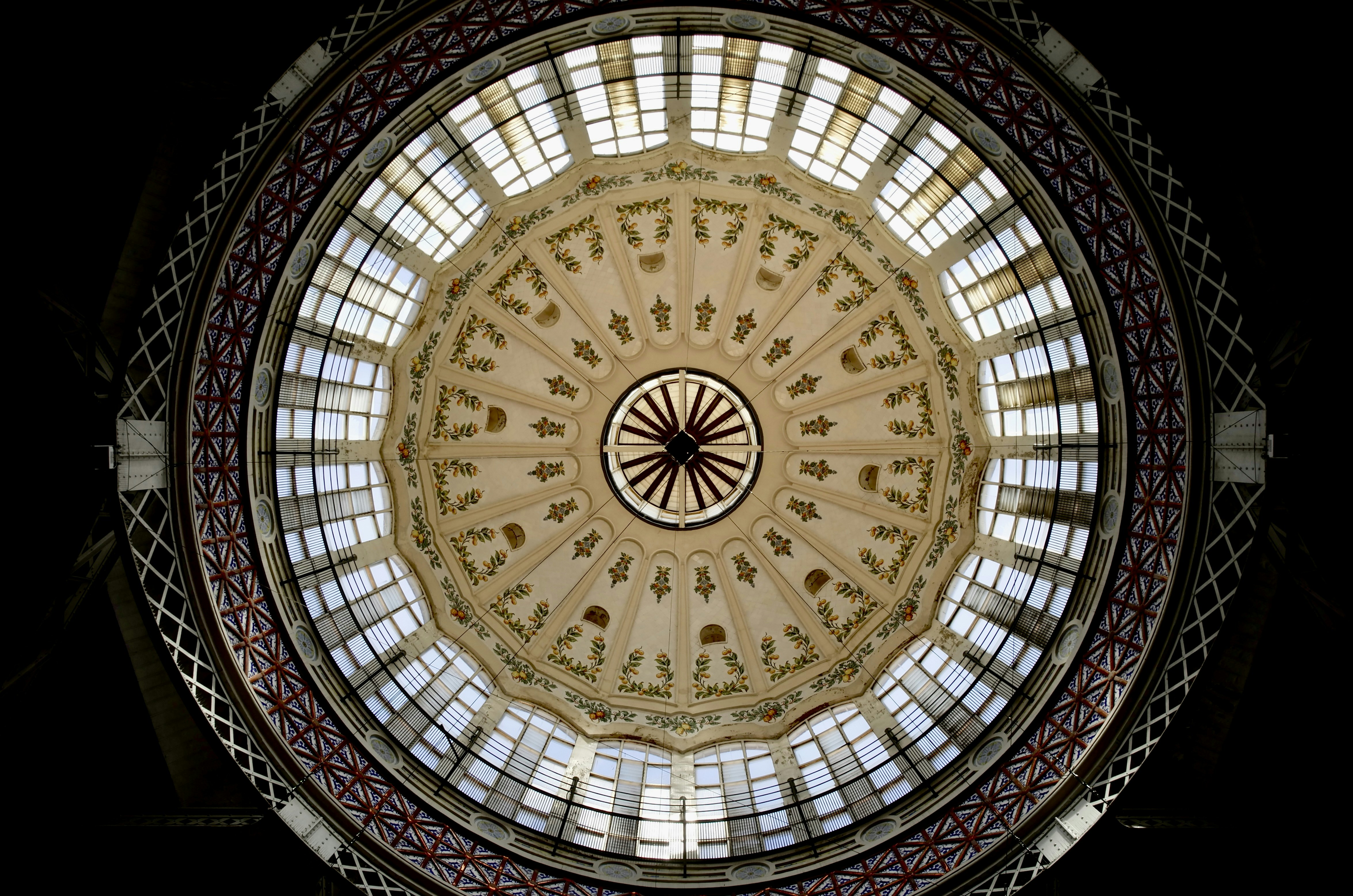 Looking Up at the Ornate Dome Ceiling of Valencia's Central Market A striking upward shot of the elaborately decorated dome ceiling of the Mercado Central in Valencia, Spain. Floral painted motifs in orange and green repeat across the vaulted panels, surrounding a central skylight that floods the space with natural light. The intricate ironwork lattice of the outer ring frames the composition in dark contrast. Shot directly overhead, the perfect circular symmetry creates an almost hypnotic, mandala-like effect that is as much fine art as it is architectural photography.