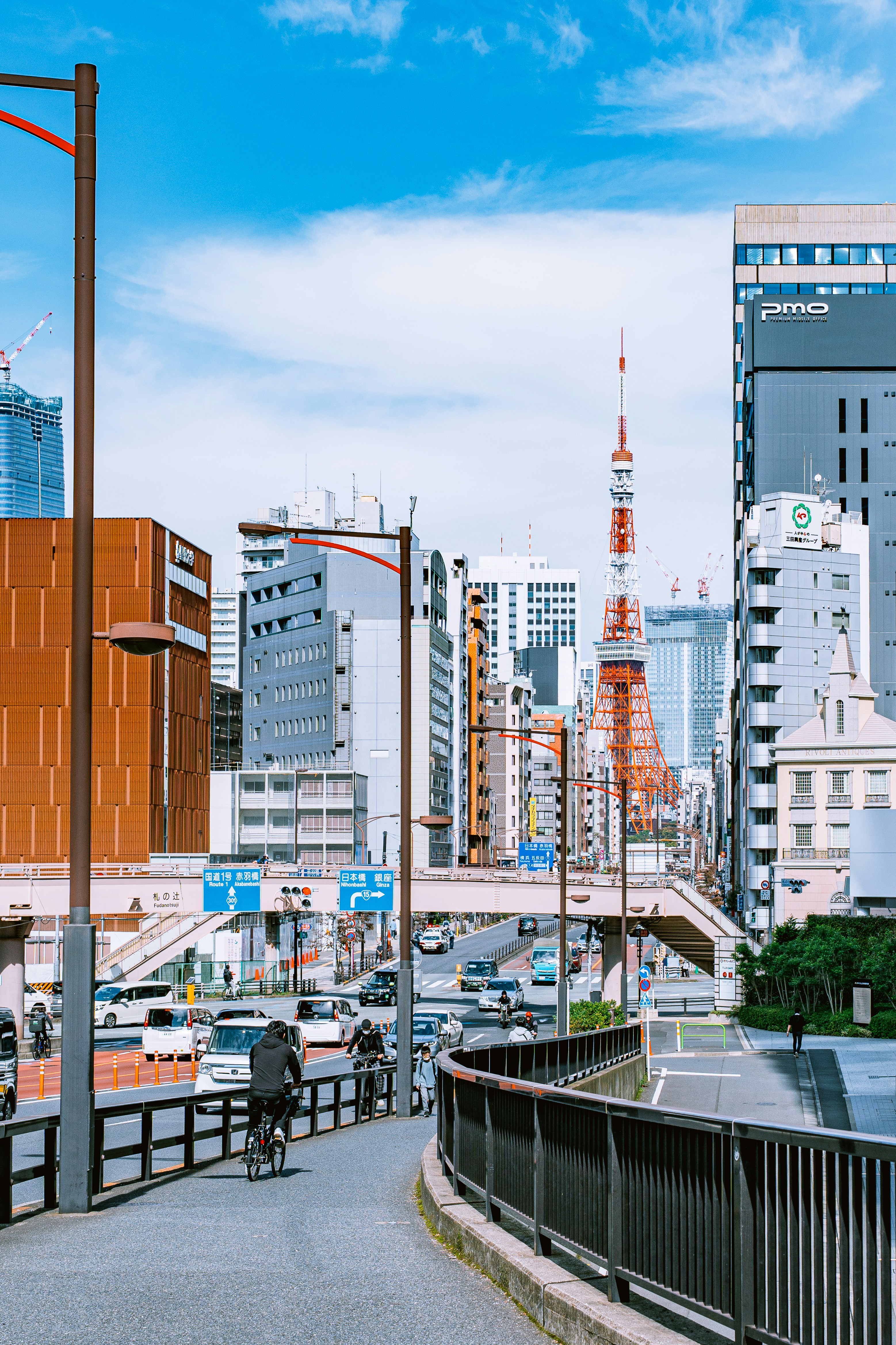 A morning cityscape along a pedestrian-friendly avenue with Tokyo Tower rising behind a cluster of modern buildings and a curving elevated walkway; the scene captures busy street life beneath a bright blue sky.