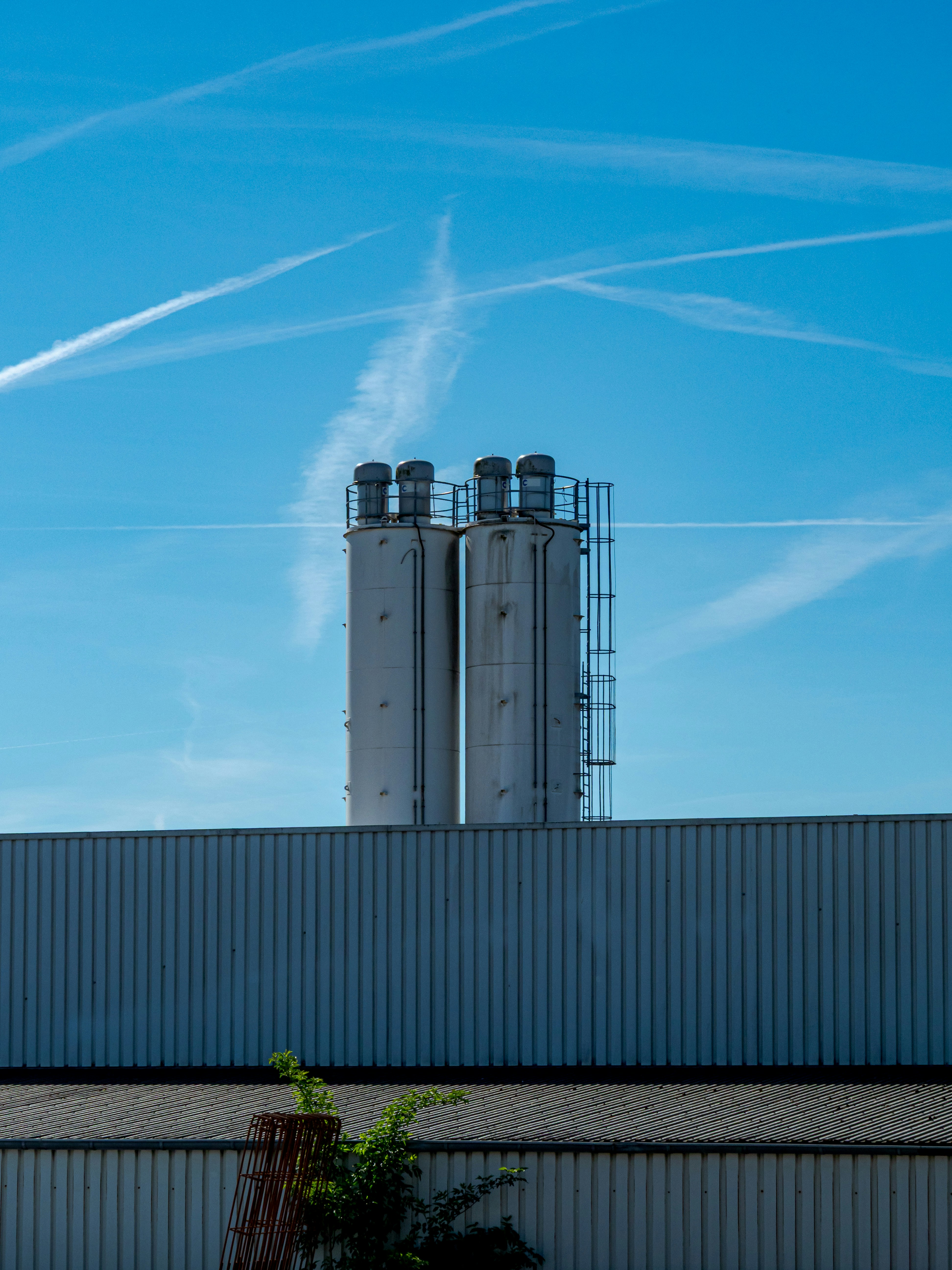 Industrial Silhouette: twin chimneys rise above a corrugated roof under a sky of contrails