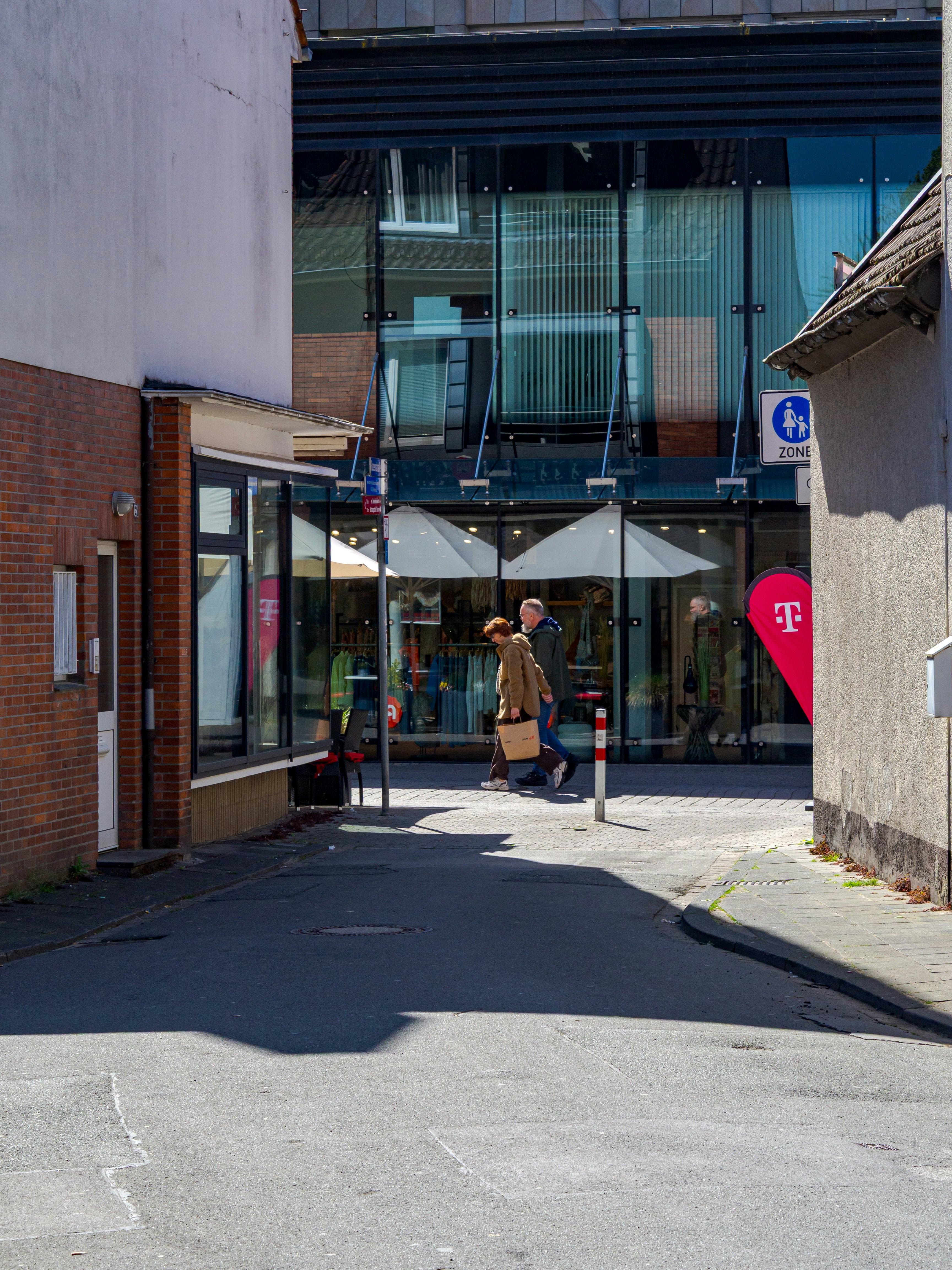 Pedestrian in a sunlit alley toward a glass-fronted shop with umbrellas