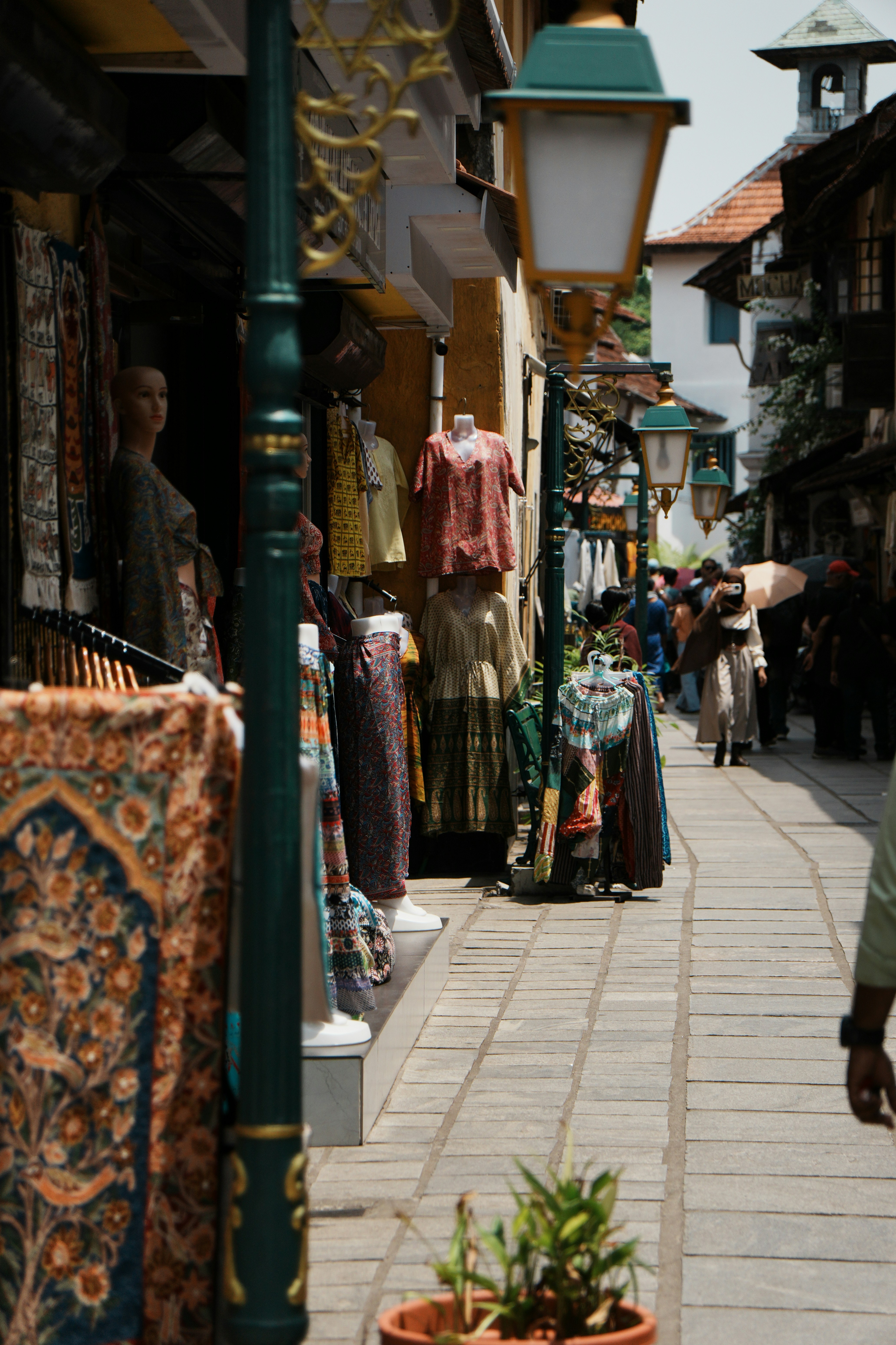 Market Stroll Under Warm Lanterns: Fabrics and passerby along a cobblestone alley