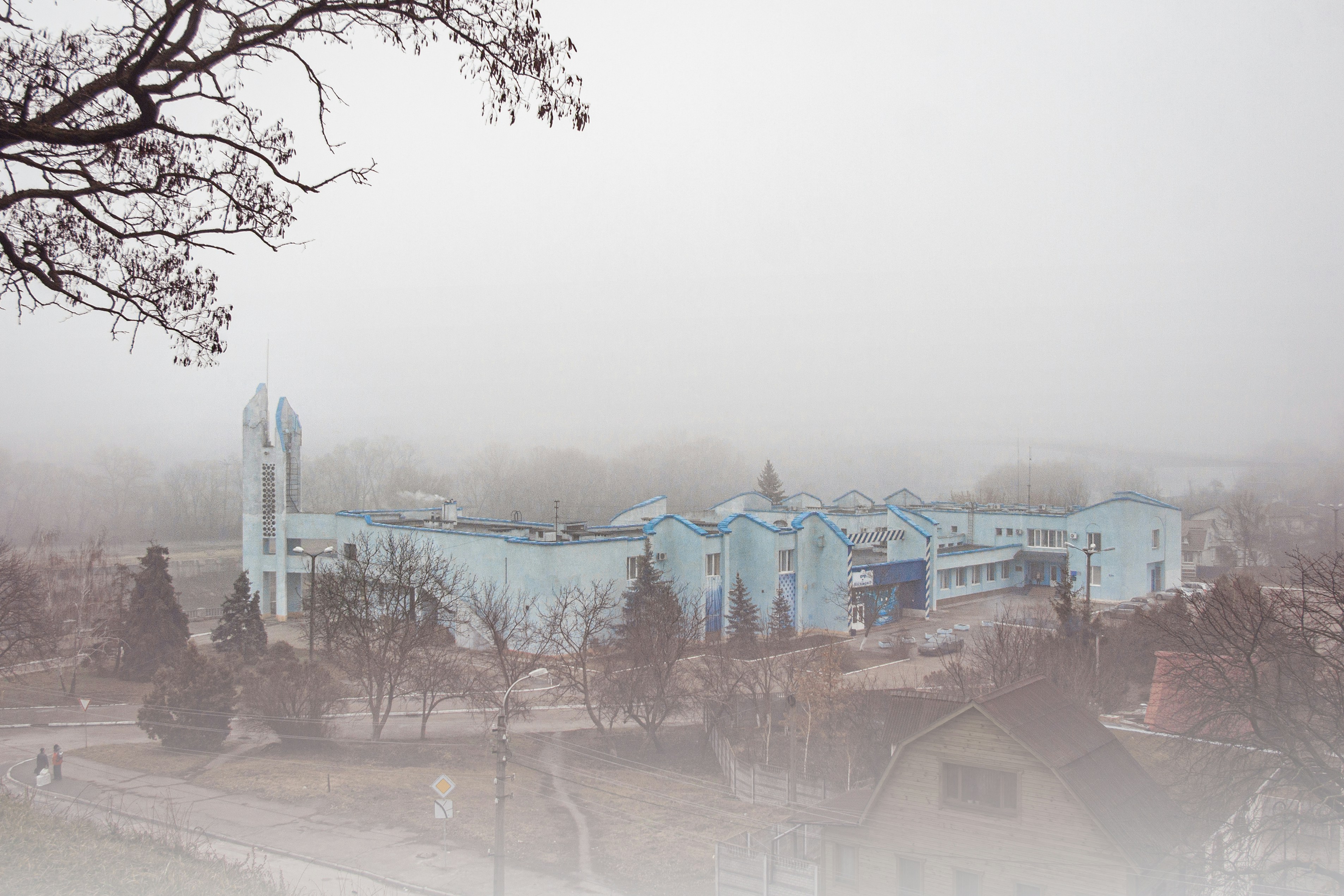 Blue building complex seen through soft morning mist with bare trees in foreground; overall hazy, high-key scene emphasizing architectural shapes and subdued tones.