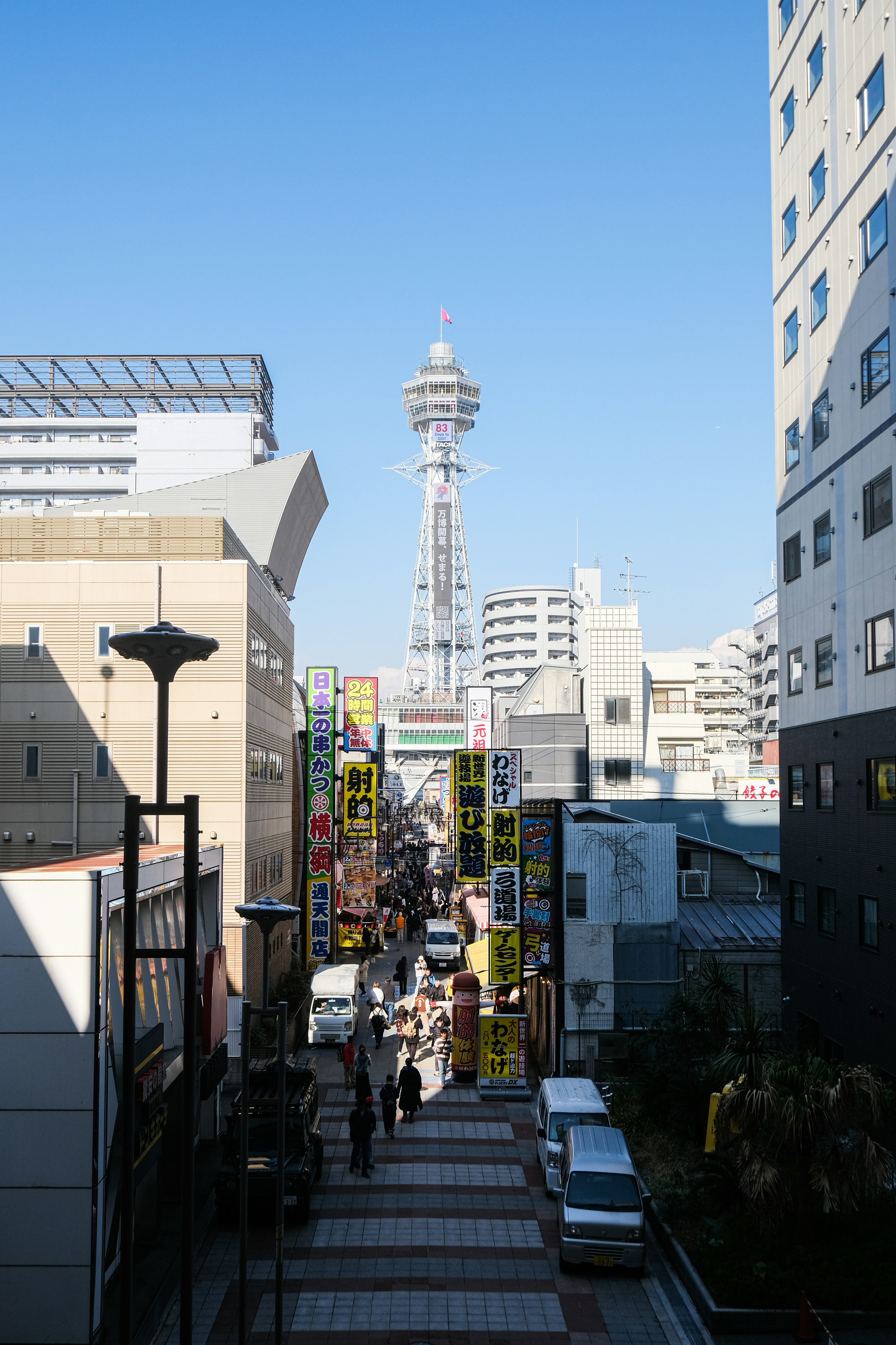Urban street scene in Japan with signs in Japanese, pedestrians, and a distant Tokyo Tower rising above mid-rise buildings; a high-angle view captures vibrant storefronts and a clear blue sky.