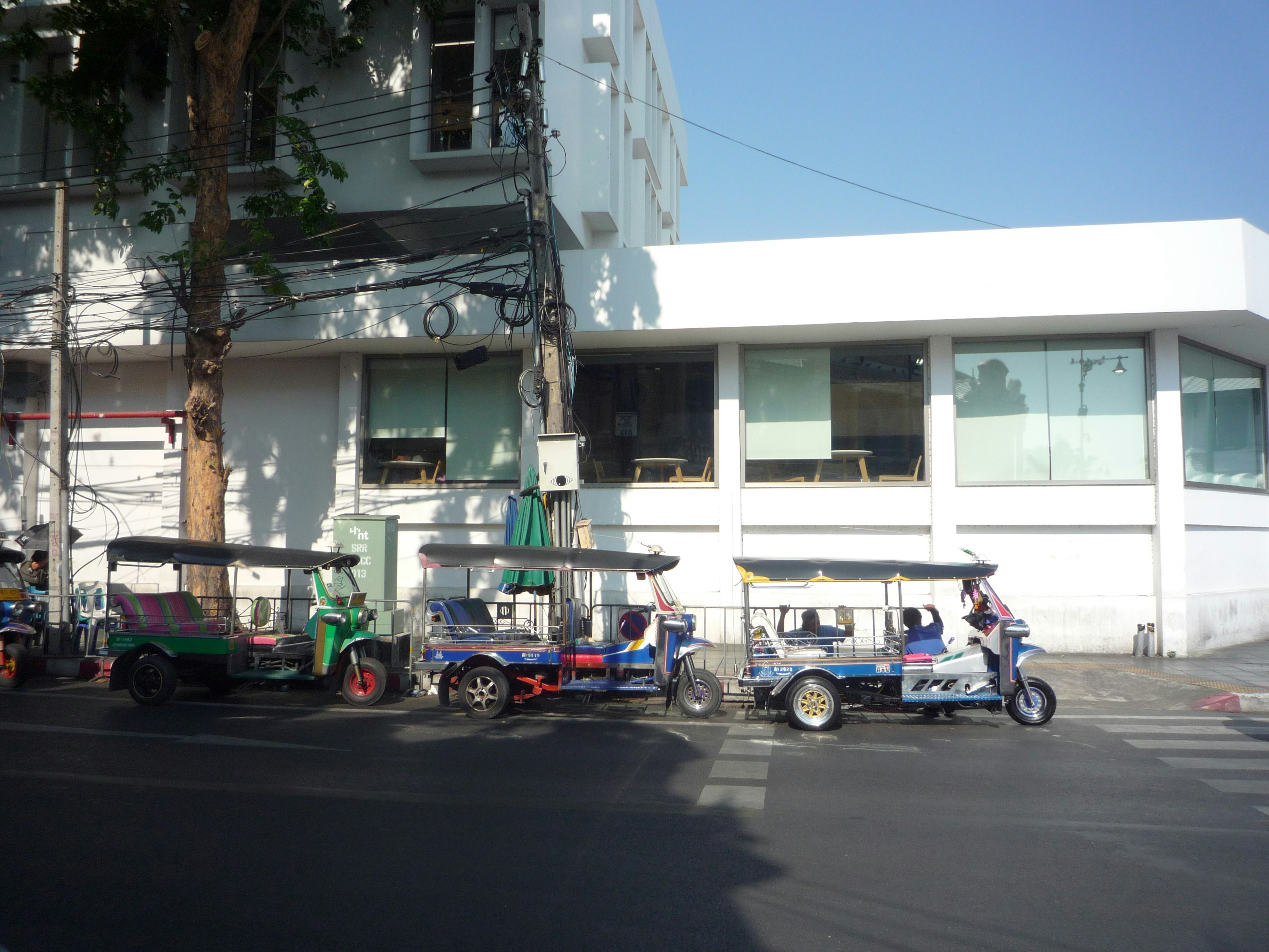 Line of colorful tuk-tuks parked along a city street in front of a white modern building with large windows.