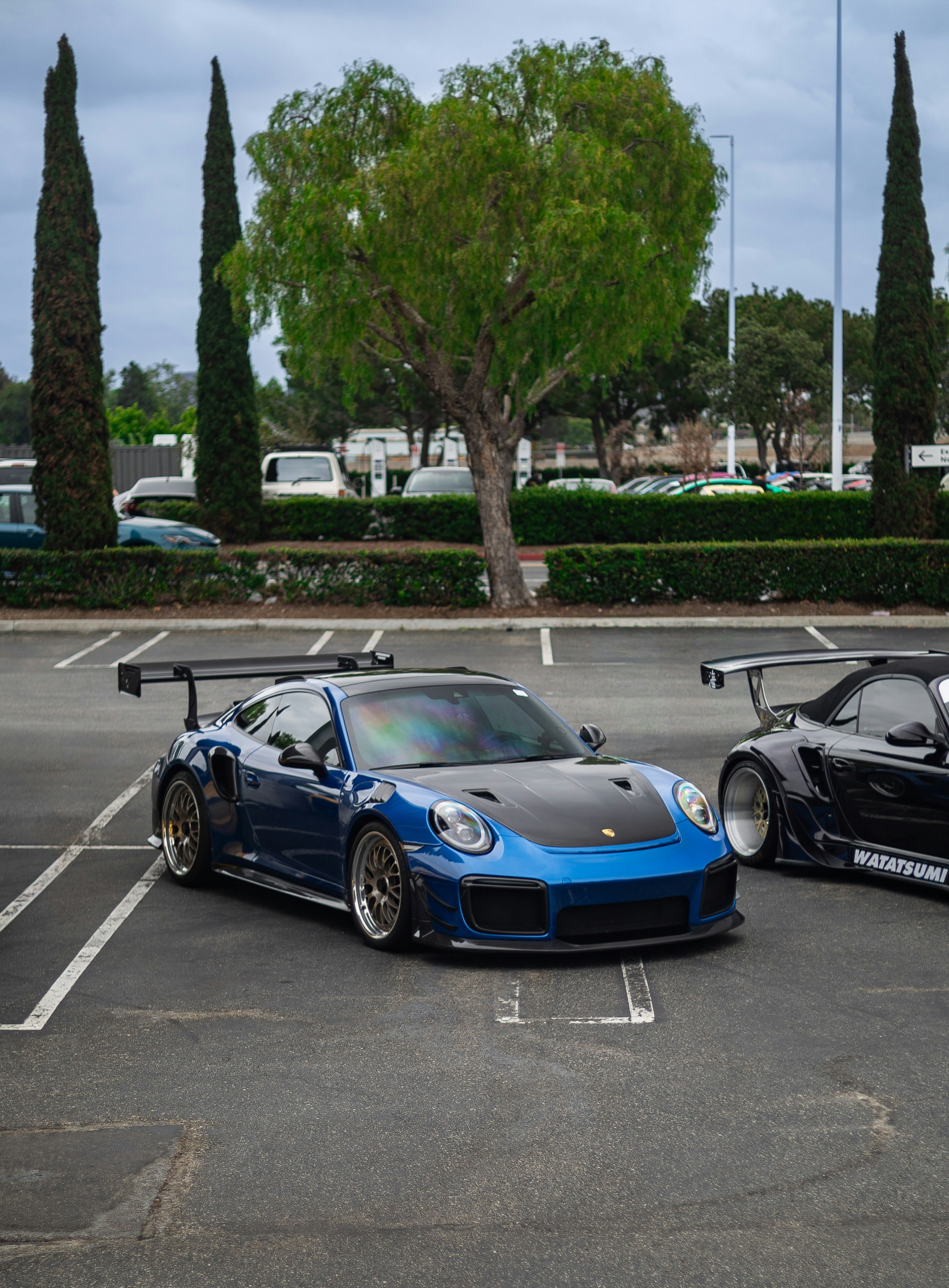 Blue Porsche race car with large rear wing parked in a lot, flanked by a black rival, green trees and a cloudy sky in the background. Car details and lines are emphasized; a showroom-like setting.