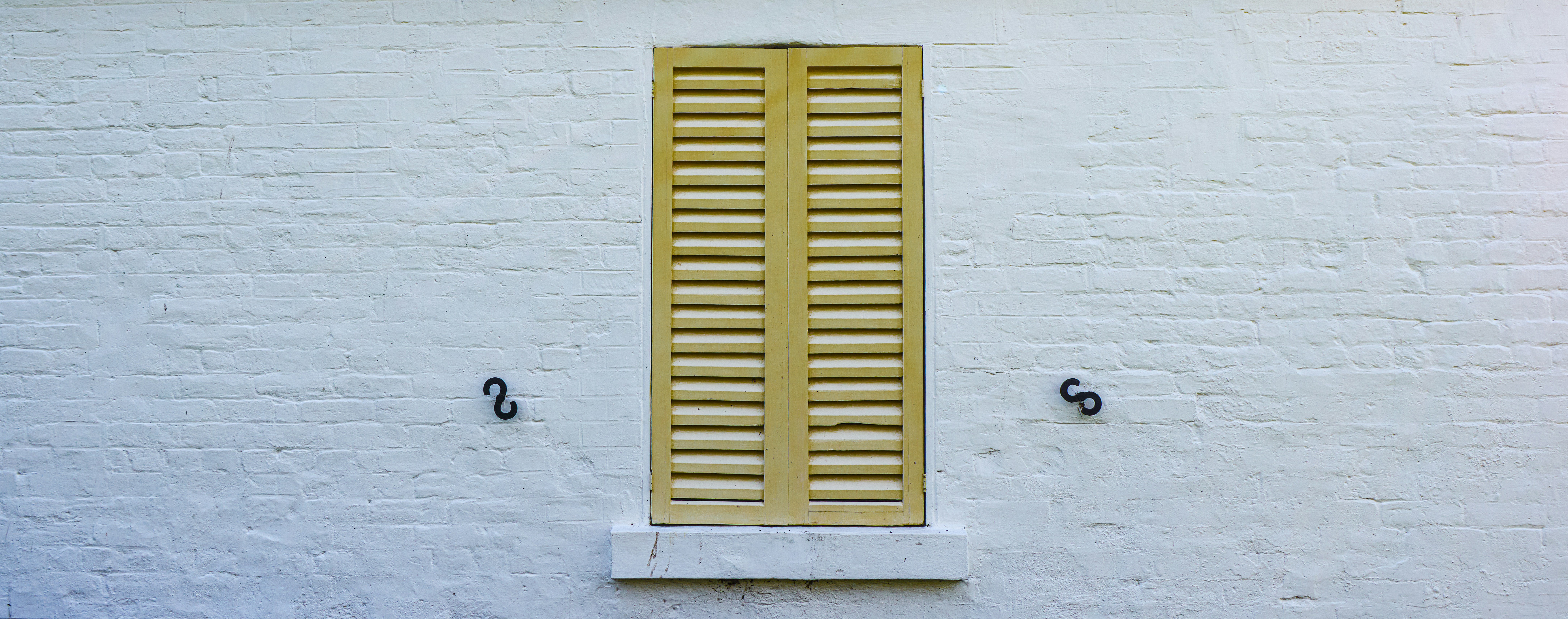 A centered golden window shutter against a pale blue brick wall, with small decorative wall hooks on either side.