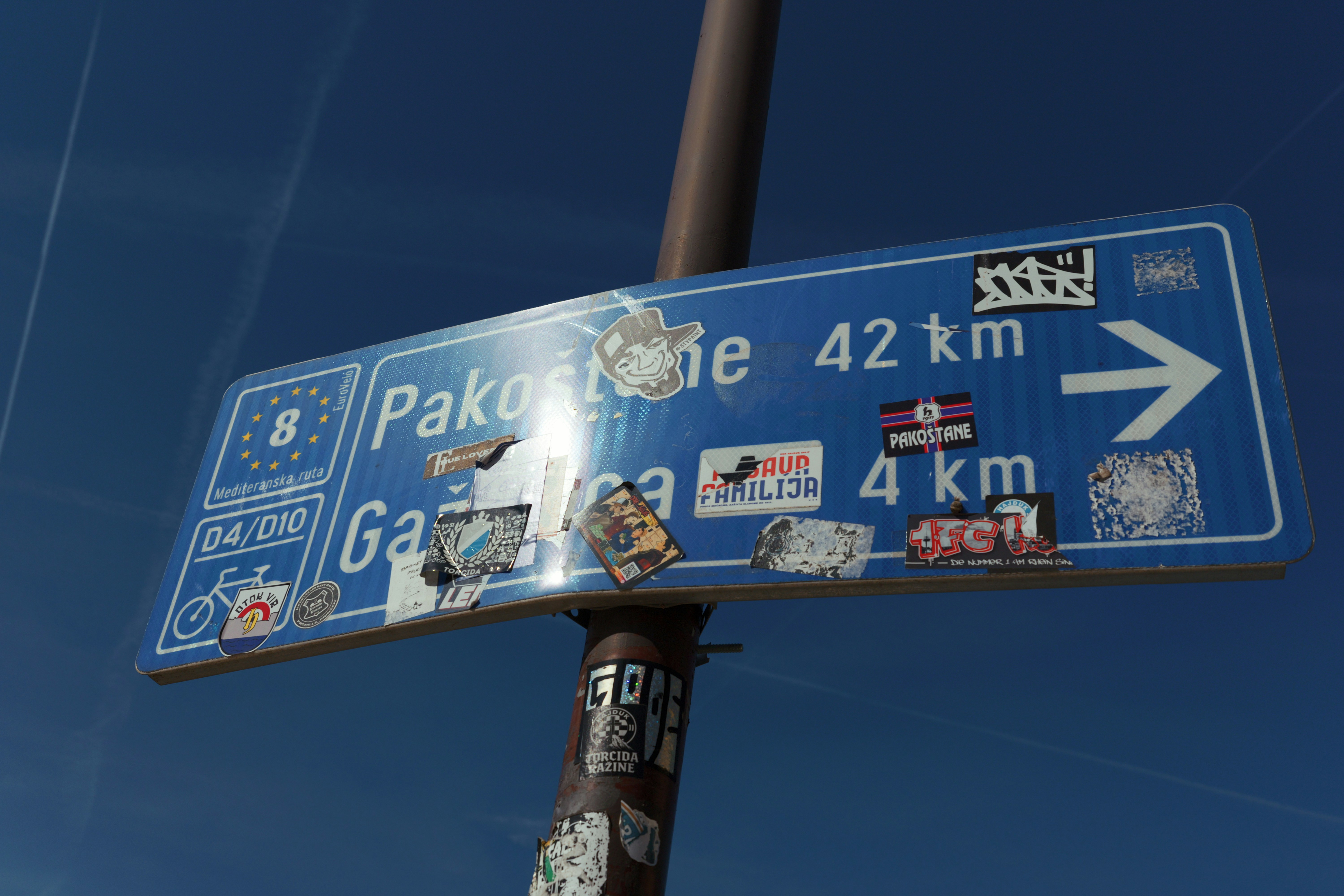 Blue road sign with distance indicators, covered in stickers, mounted on a pole against a bright blue sky.