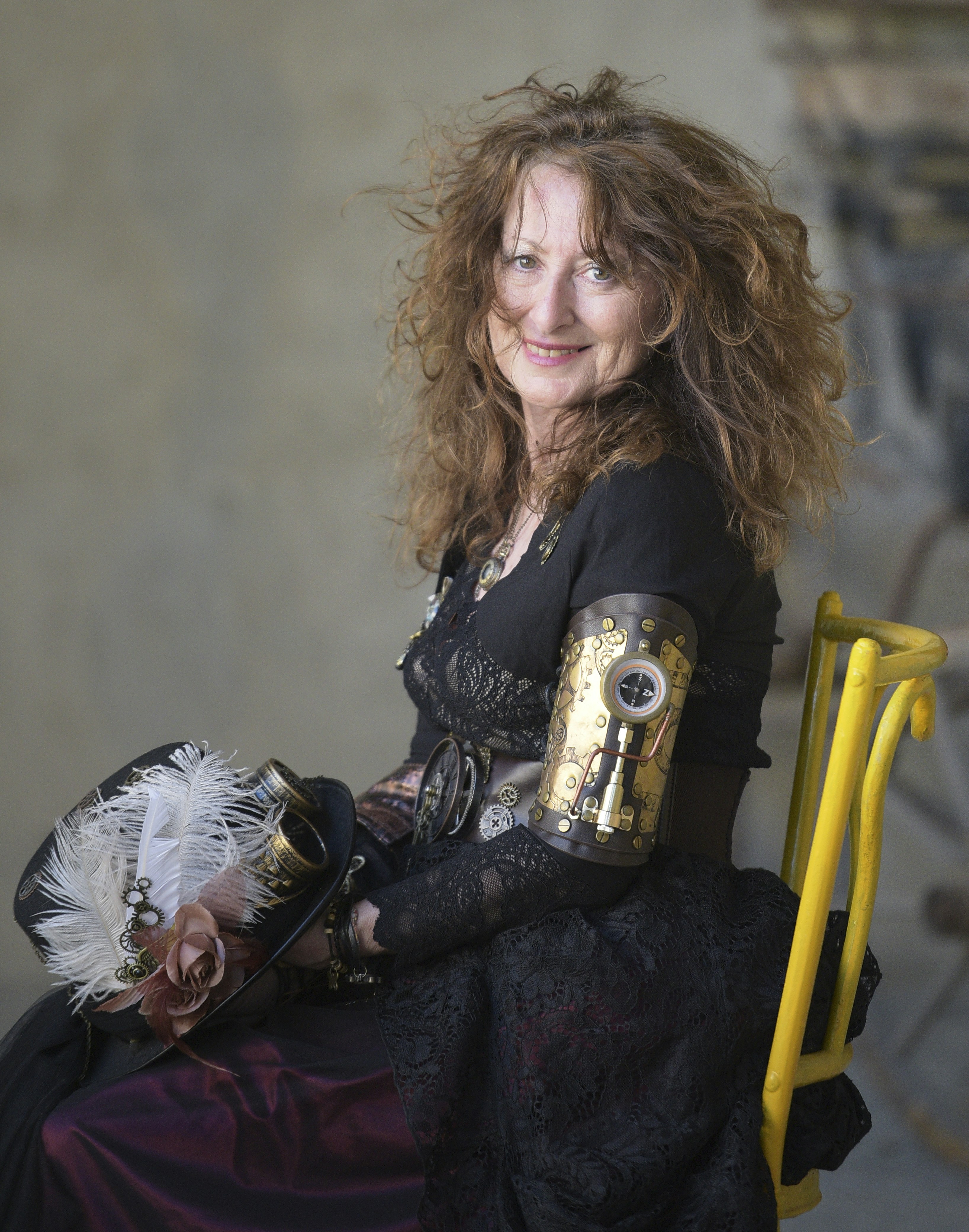 Portrait of a smiling woman in a black lace dress, wearing a retro prosthetic arm and holding a feathered hat, seated on a yellow chair against a muted backdrop. The image emphasizes character and vintage style.