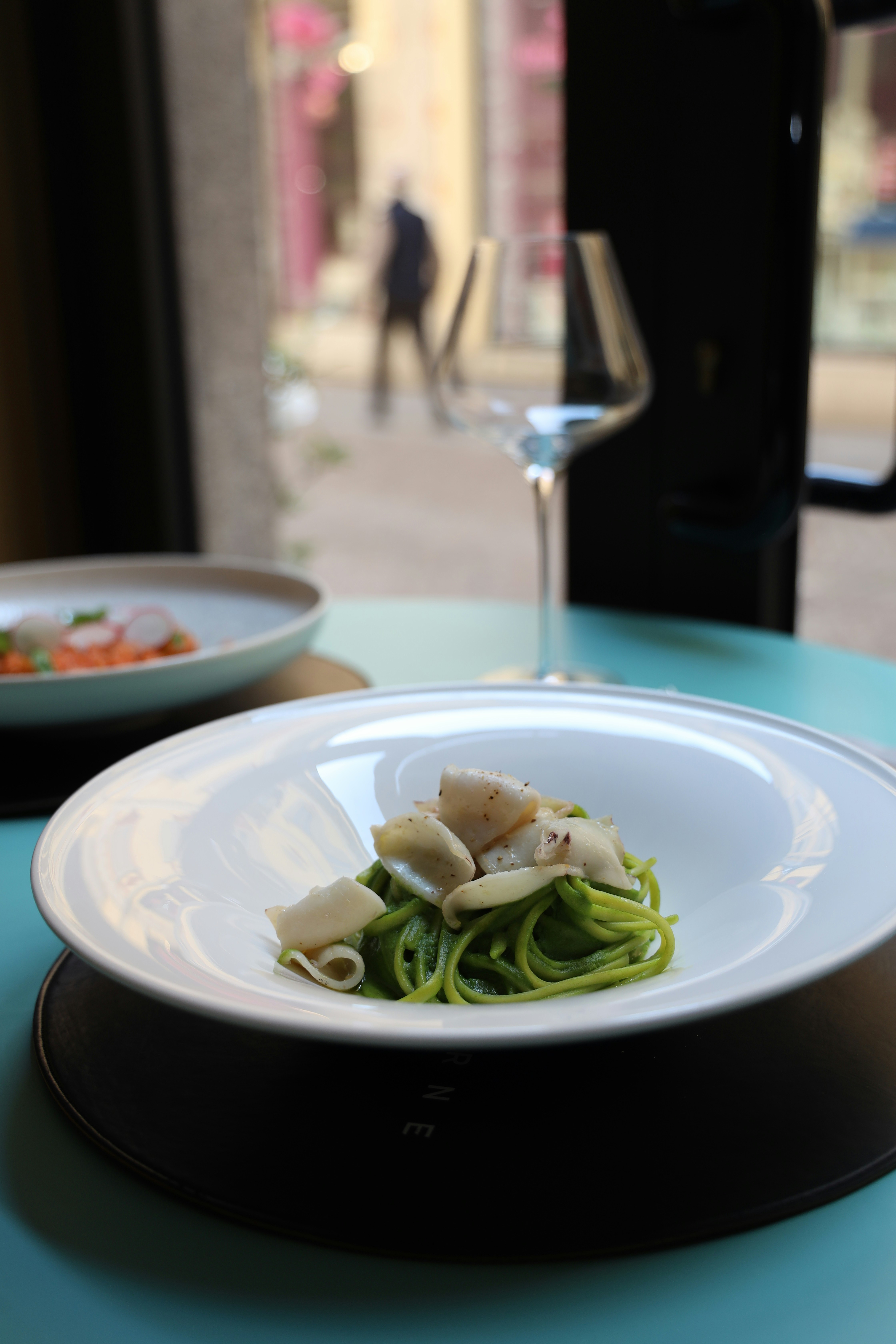 Green pasta under soft cafe light, with blurred street scene beyond