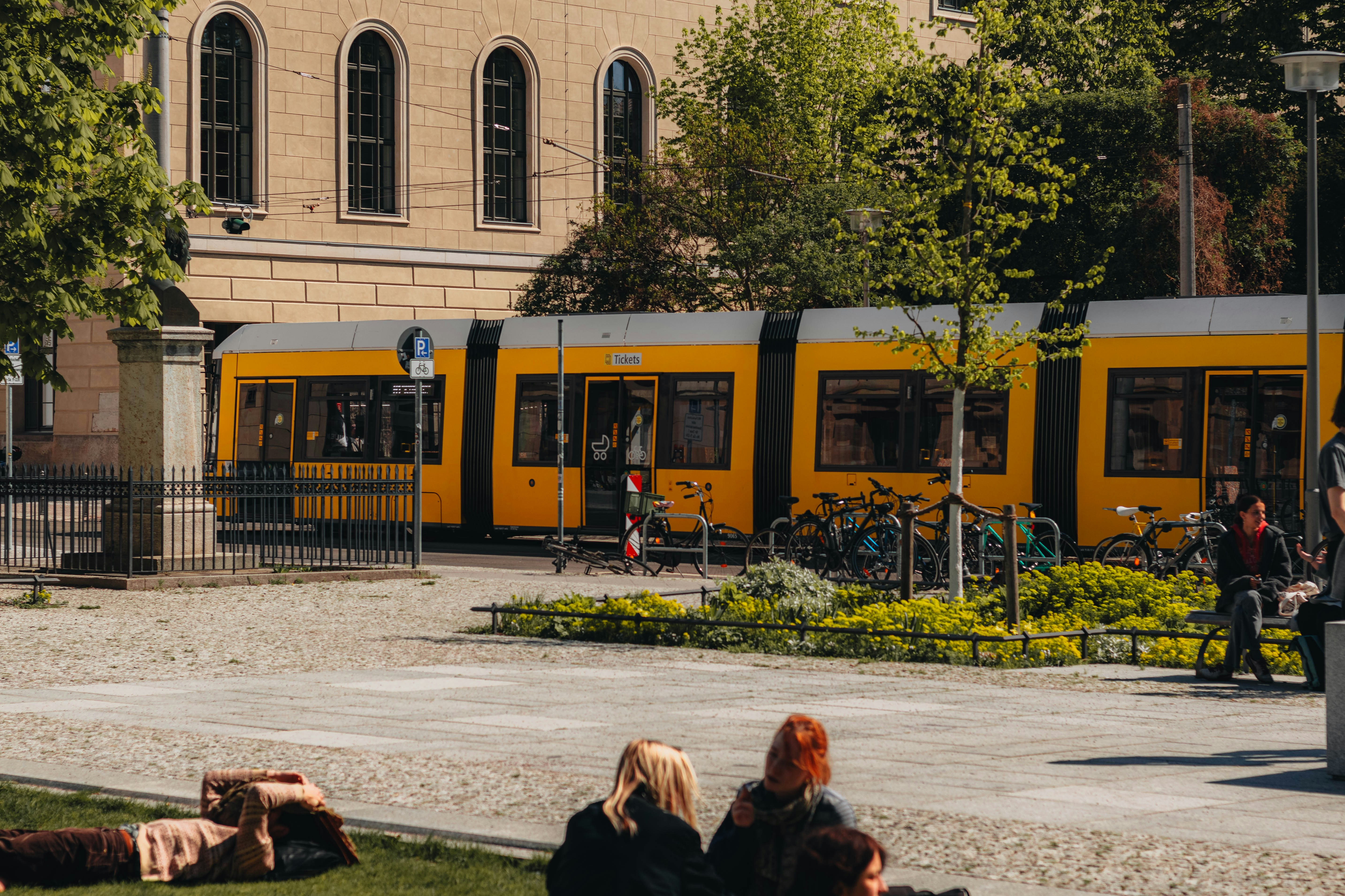 A yellow tram stops near a park with people.