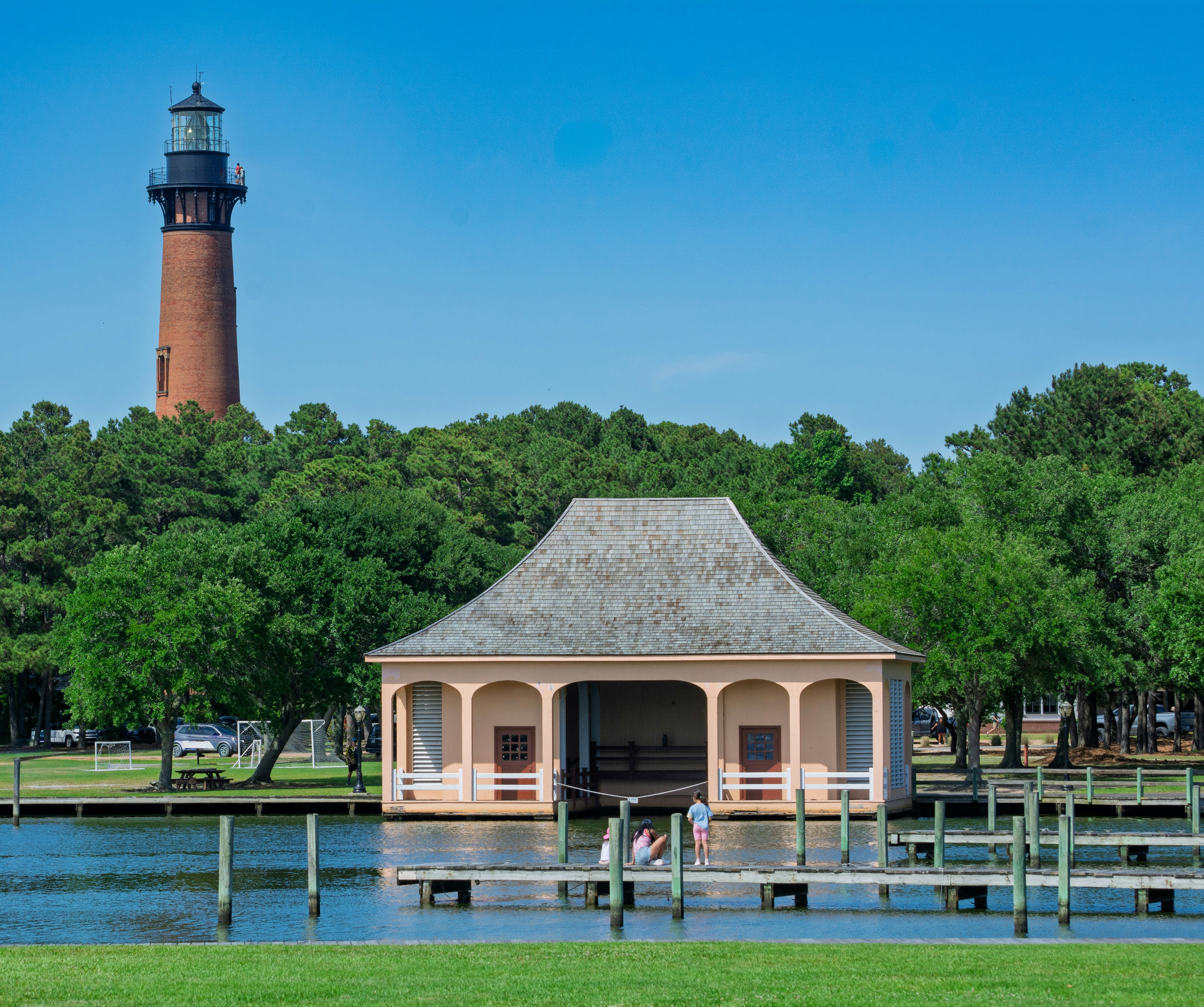 Lighthouse rises behind a calm waterfront pavilion with a wooden dock, green trees, and a clear blue sky.