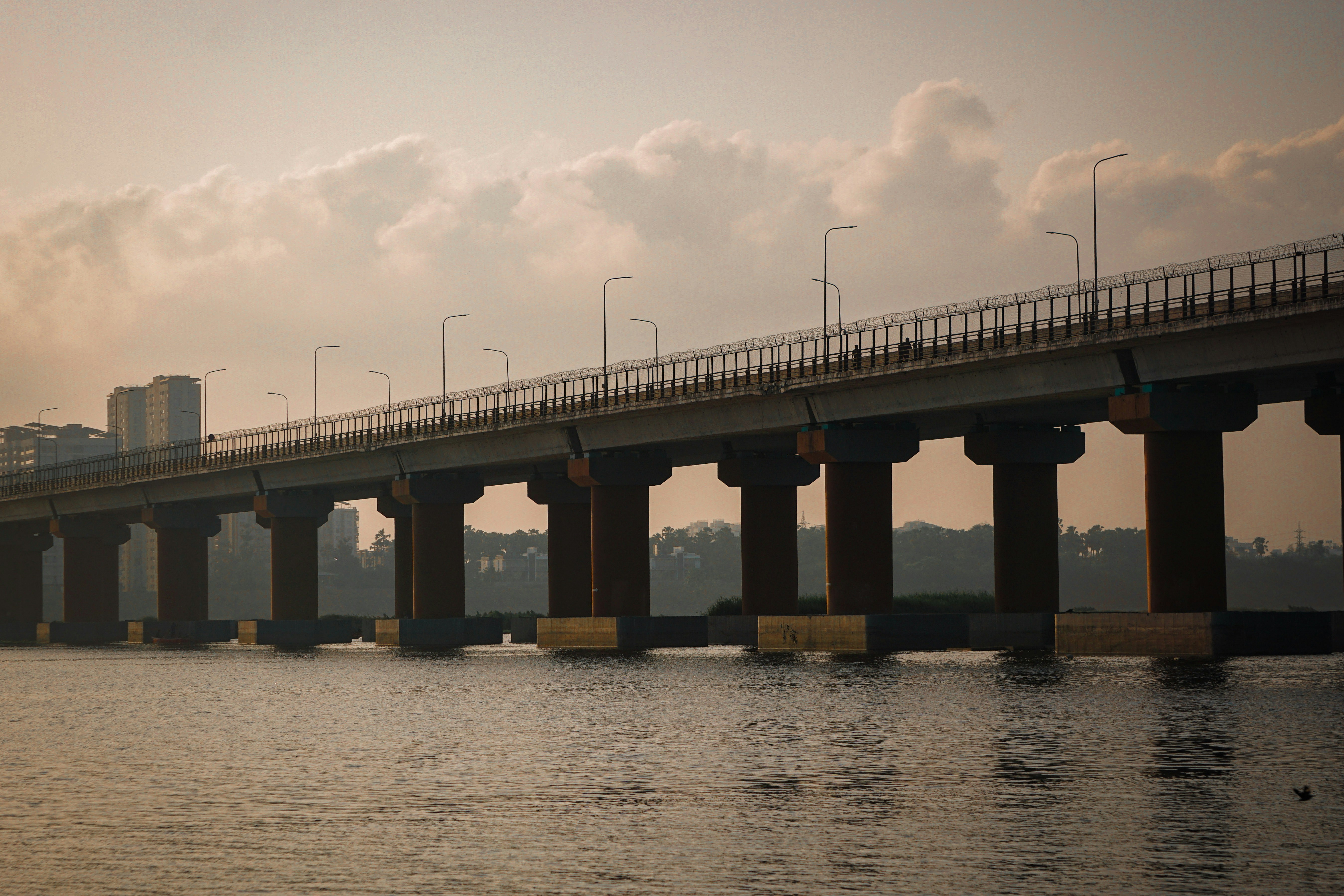 Bridge spanning a calm river at golden hour with soft clouds and distant city silhouettes in the background.