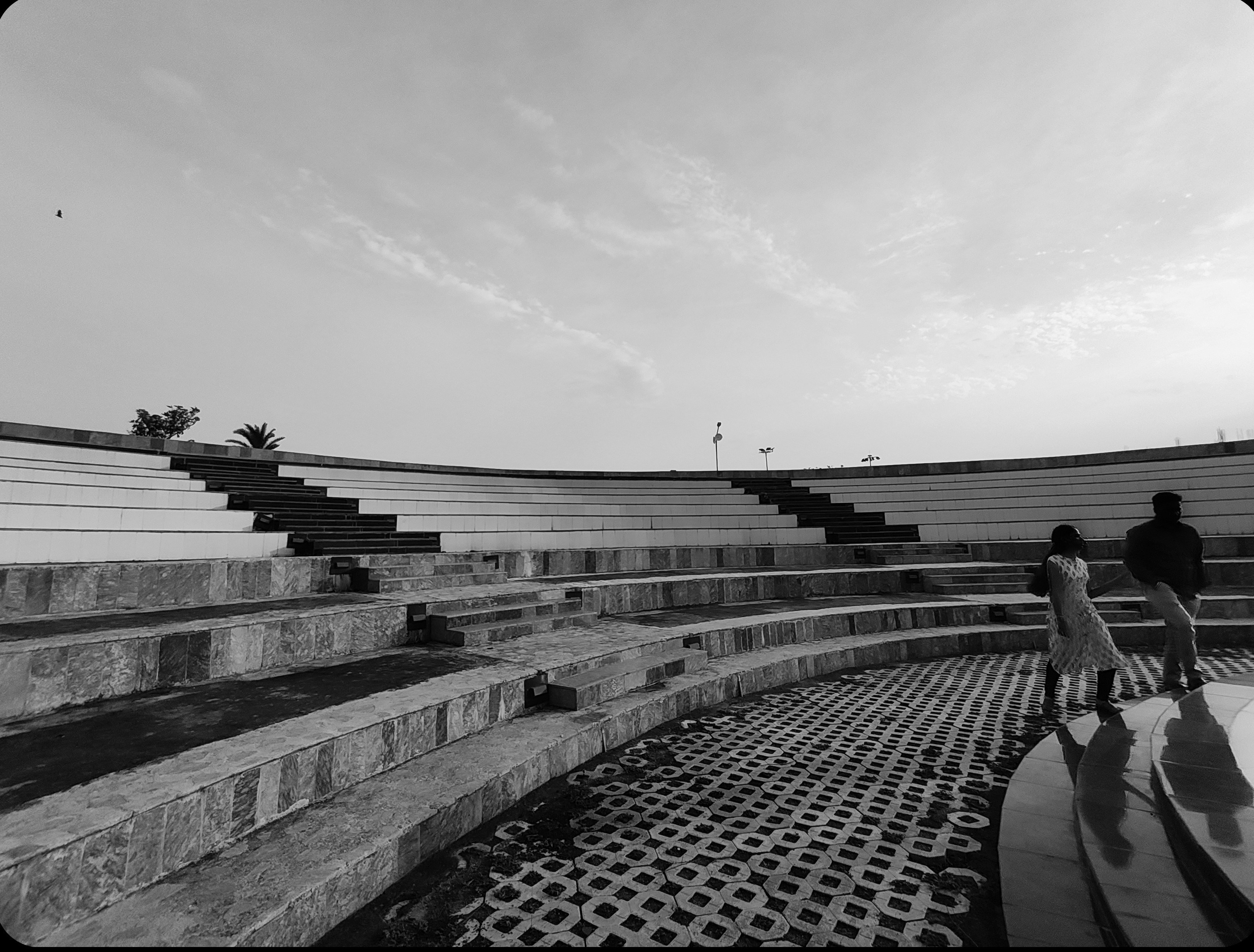 Monochrome amphitheater steps with two figures walking near the edge, curved seating and textured ground patterns.