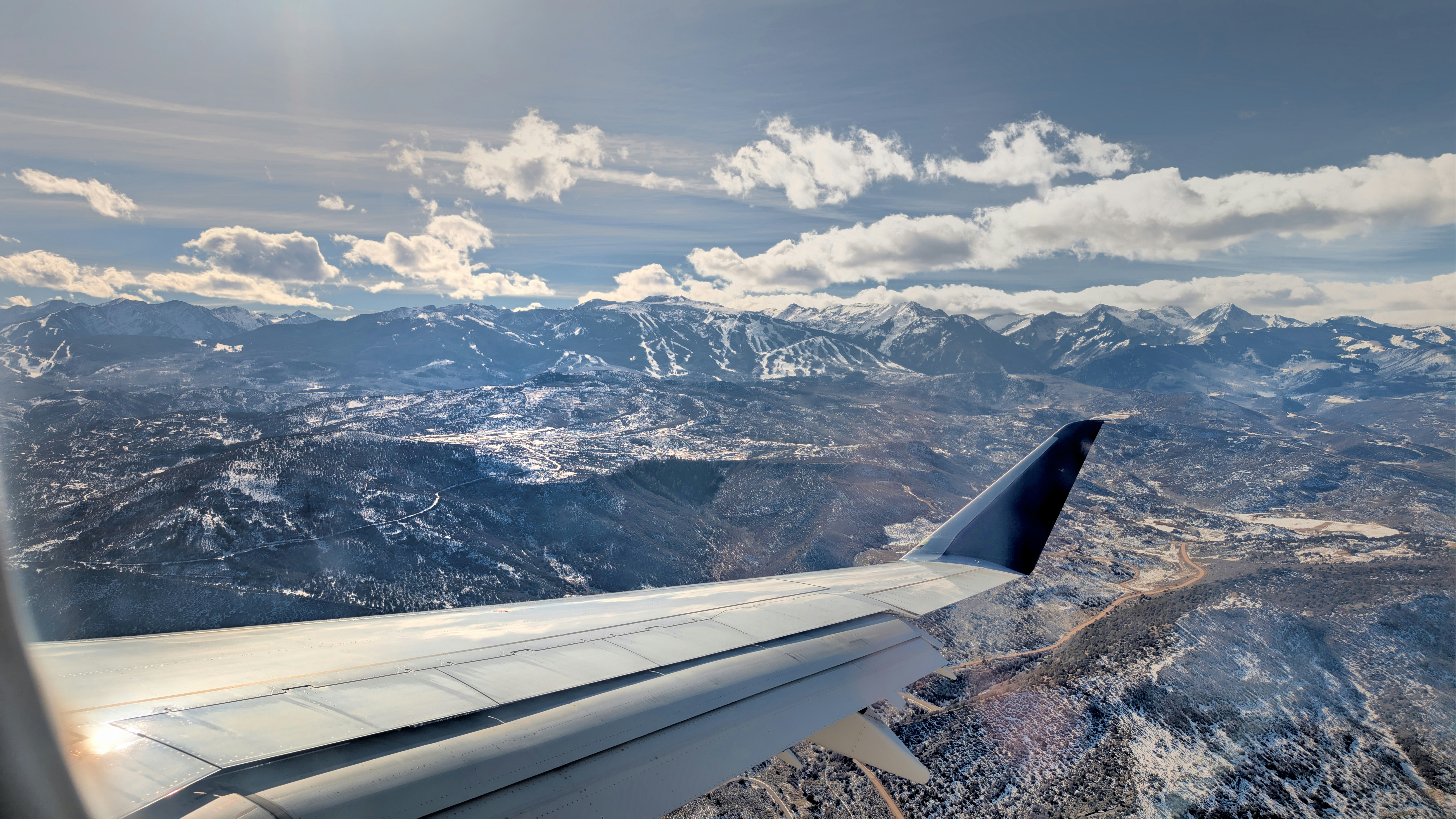 Snow-dusted mountains roll beneath a metallic airplane wing, sunlight scattering across a clear sky.