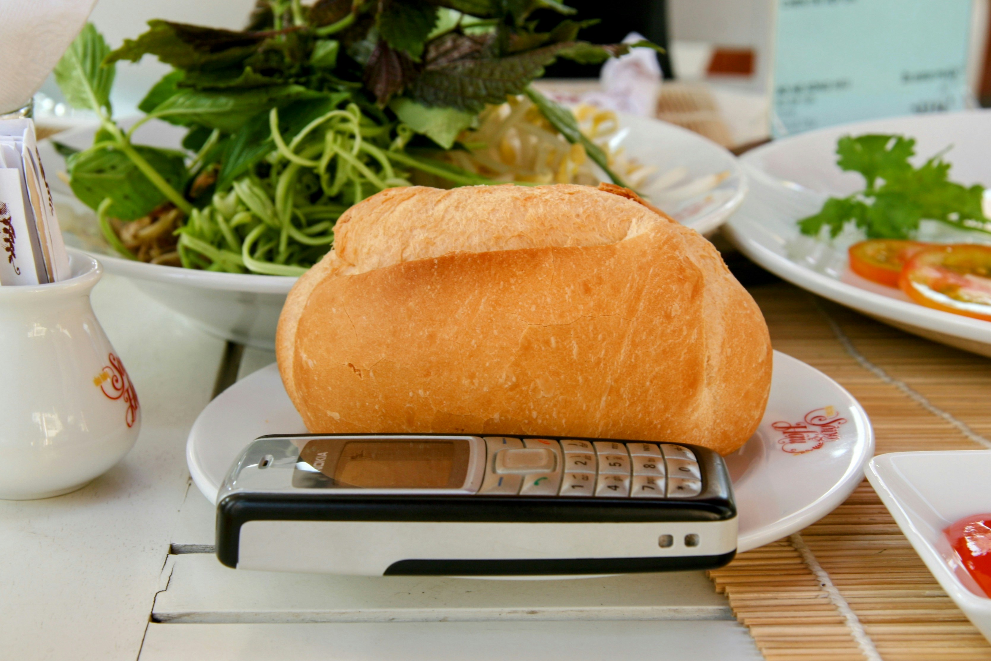 A loaf sits atop a vintage mobile phone on a white plate, surrounded by fresh greens and plated dishes.