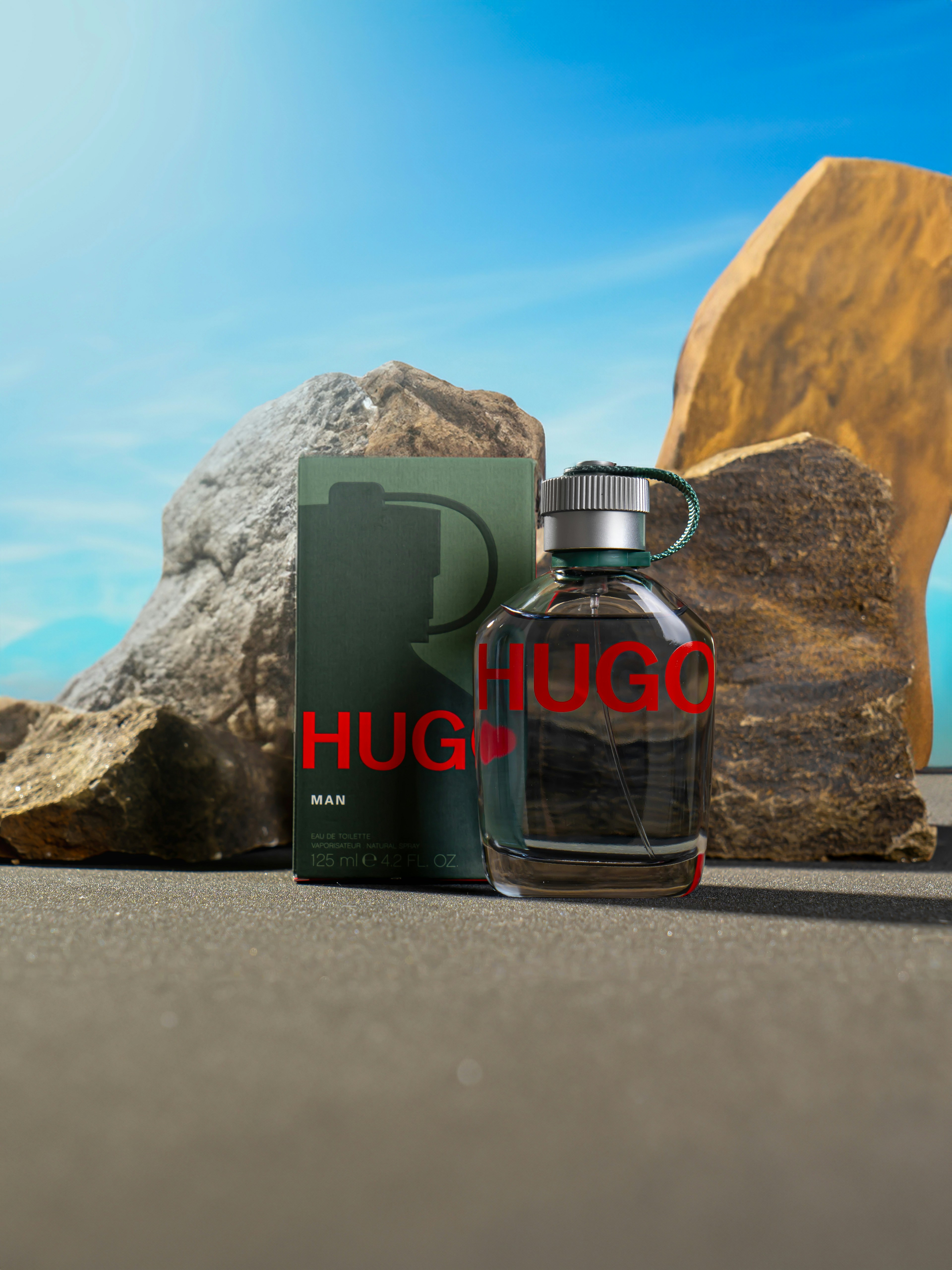A Hugo bottle and its box rest on a rocky beach setup with a blurred blue sky background. This image showcases the perfume in a minimal still-life composition, emphasizing the branding and glass texture.