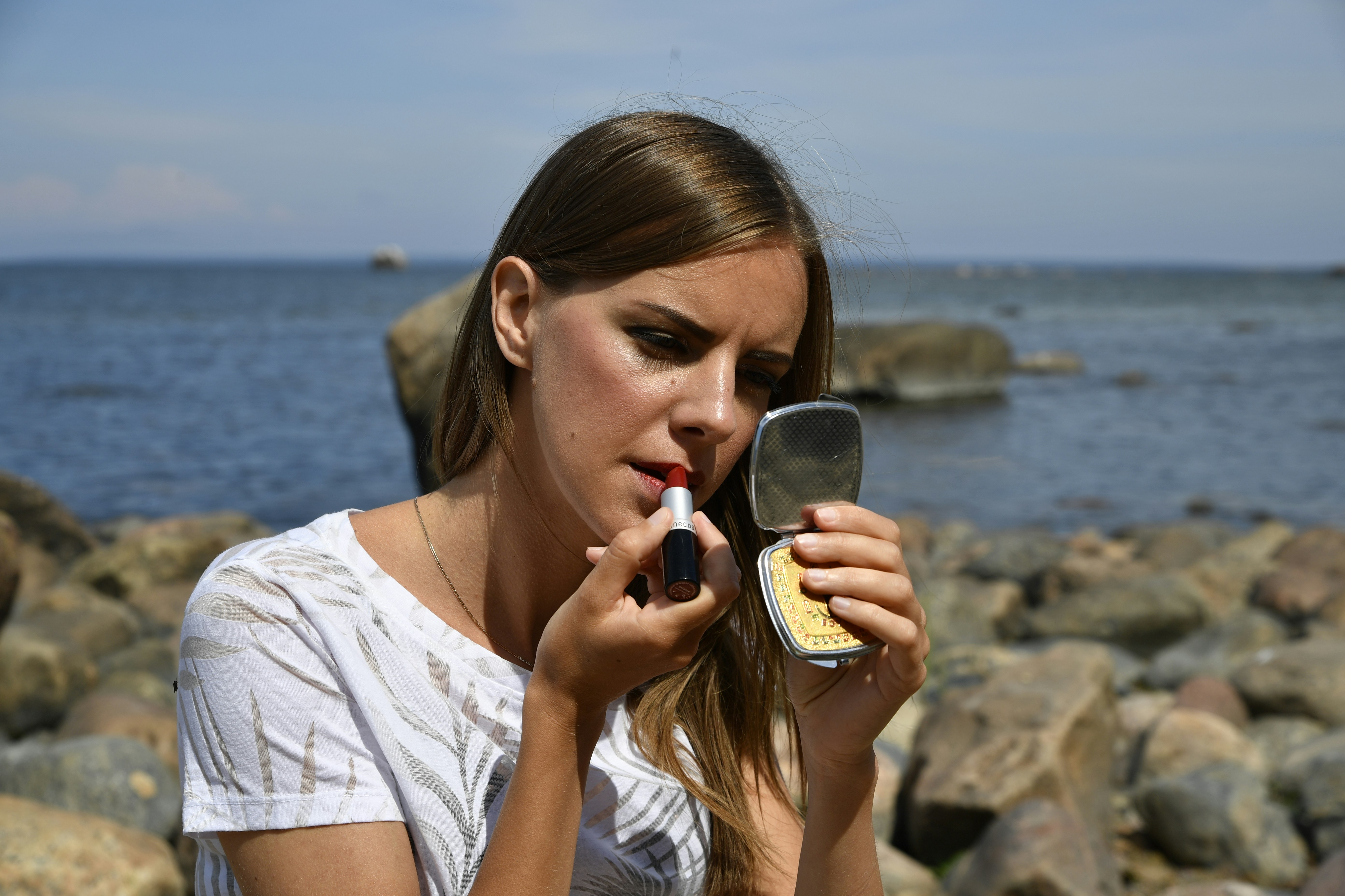 A young woman applies lipstick while checking a compact mirror beside a rocky shoreline, with sea and sky in the background. Logo/brand not visible; natural daytime lighting.