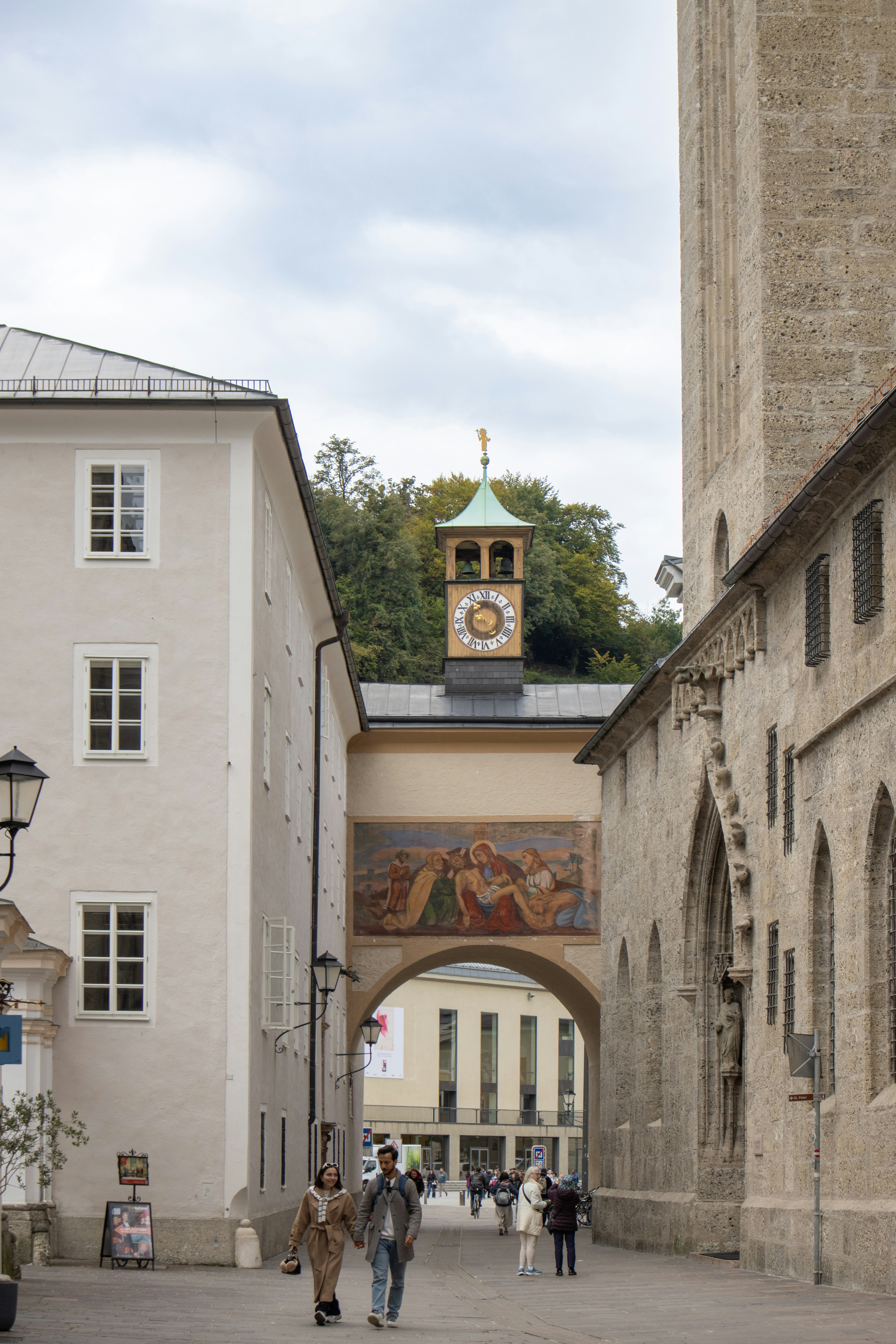 Archway between light-colored facades with a clock tower and a fresco