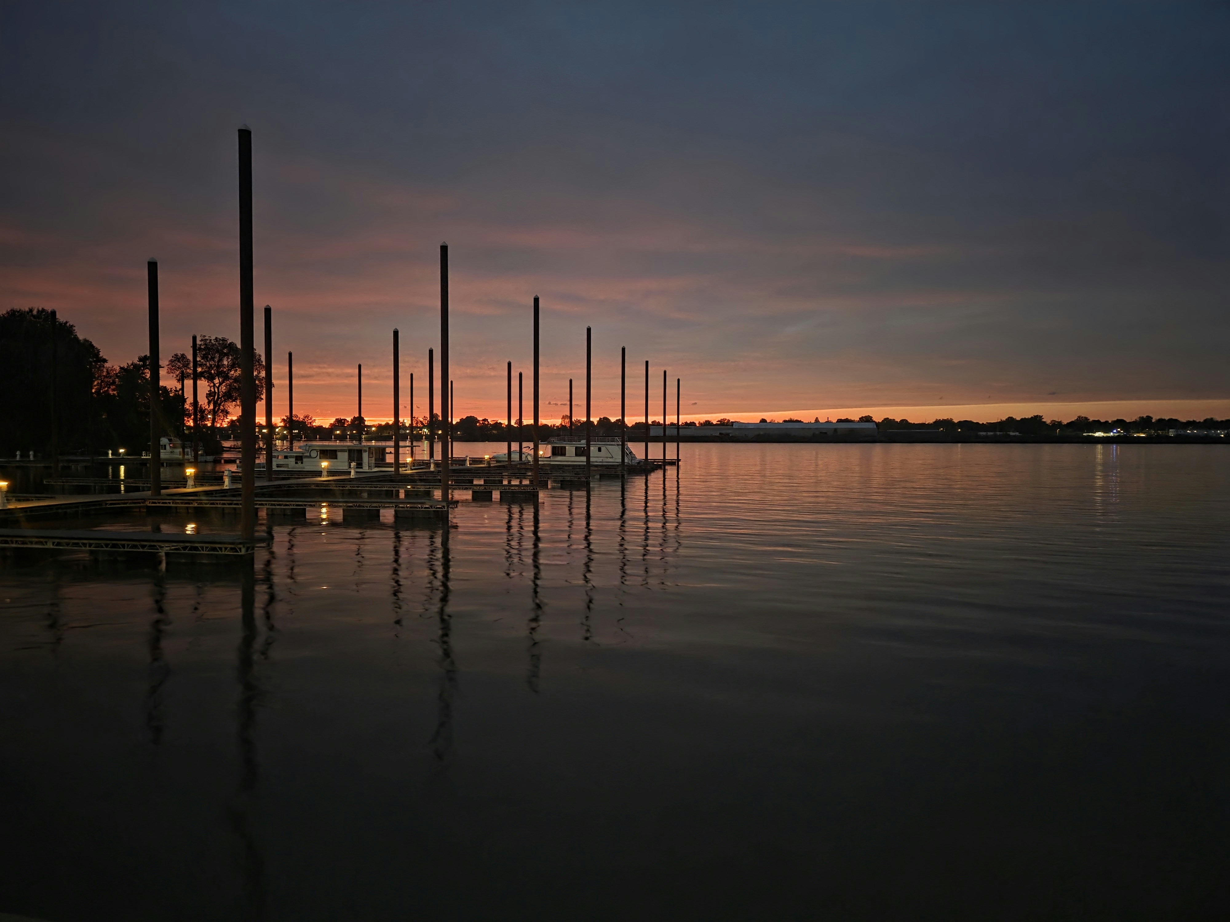Soft sunset hues reflect across calm water near a quiet marina, with vertical posts casting dark silhouettes against a gradient sky.