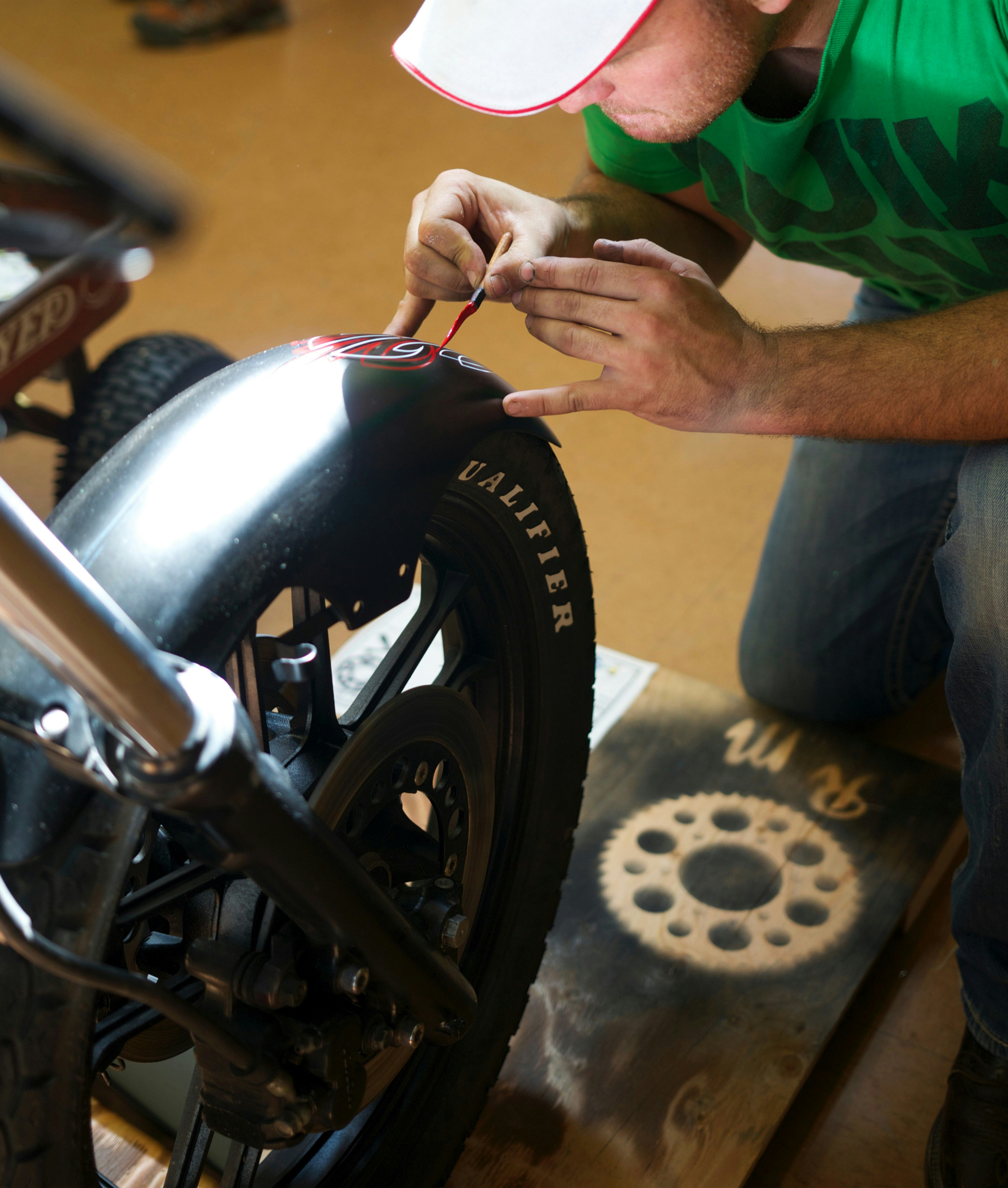 Close-up of a person kneeling beside a motorcycle wheel, hand-tooling a spoke or valve near the front tire in a workshop; tactile detail and motion suggesting a maintenance or customization task. The scene includes the tire, wheel, and surrounding bike components.