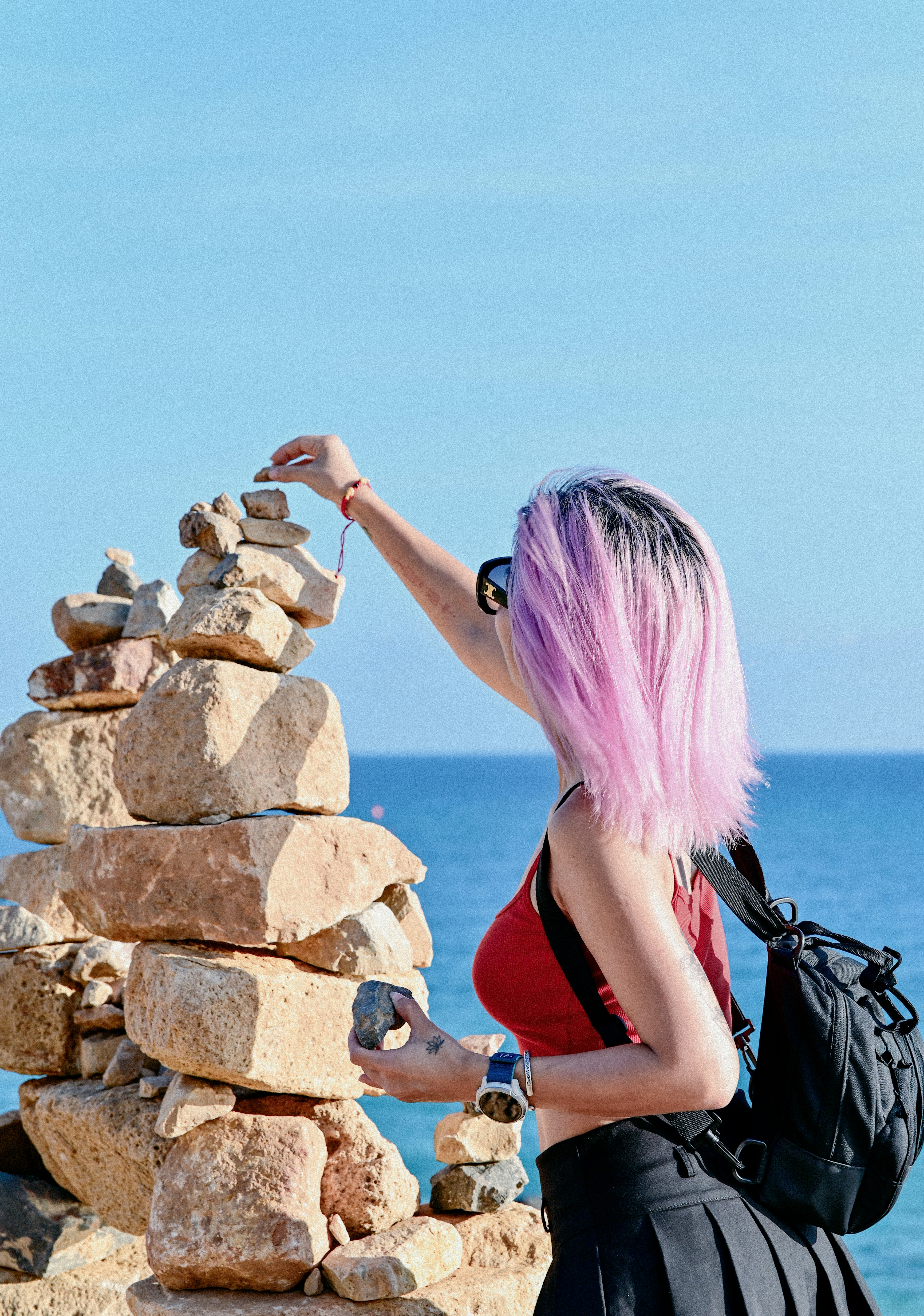 Pastel-haired explorer stacks a coastal cairn against a clear blue horizon