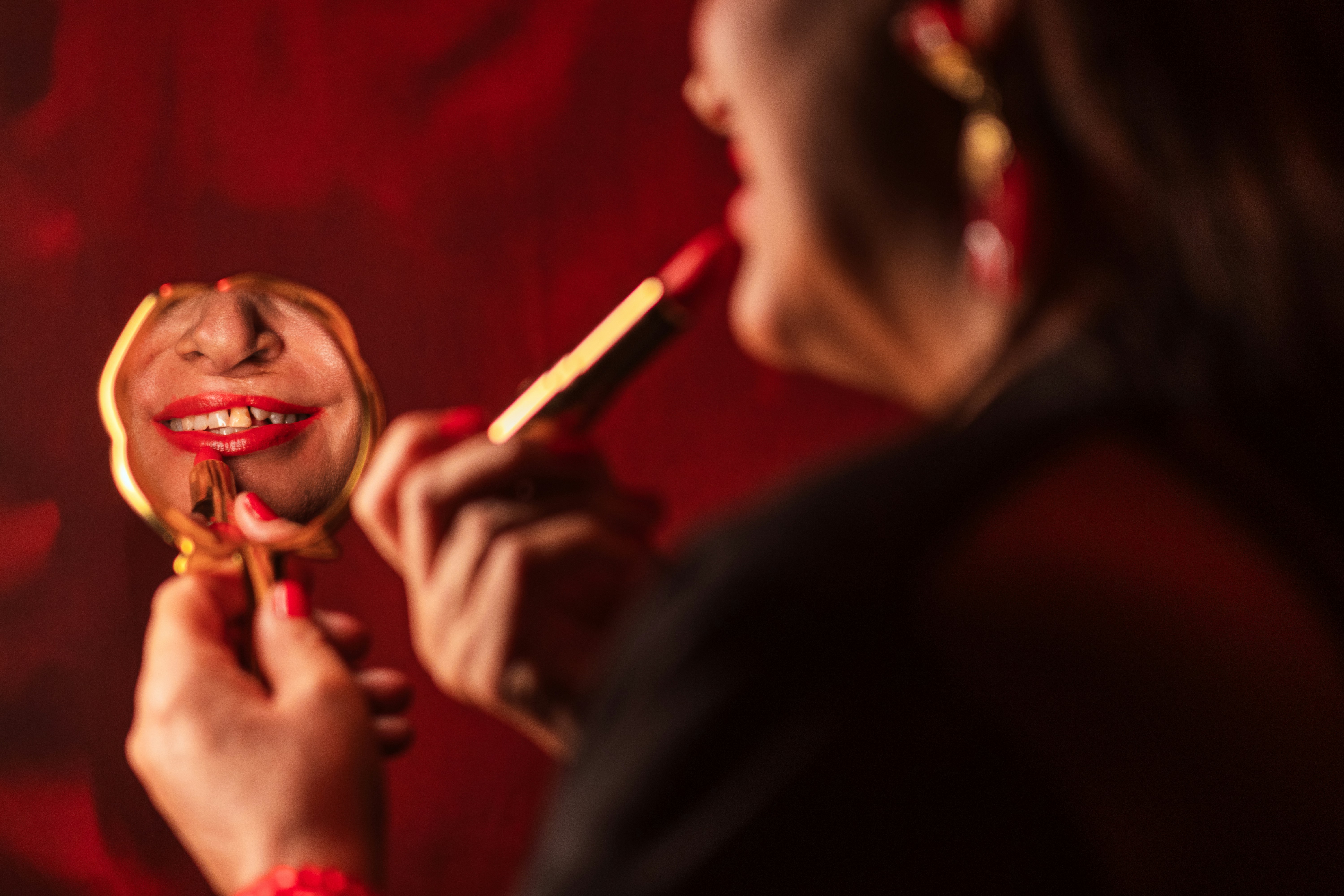A performer applies lipstick while looking into a handheld mirror, glow from red stage lighting reflecting on the face. This is a candid capture of makeup application during a performance.