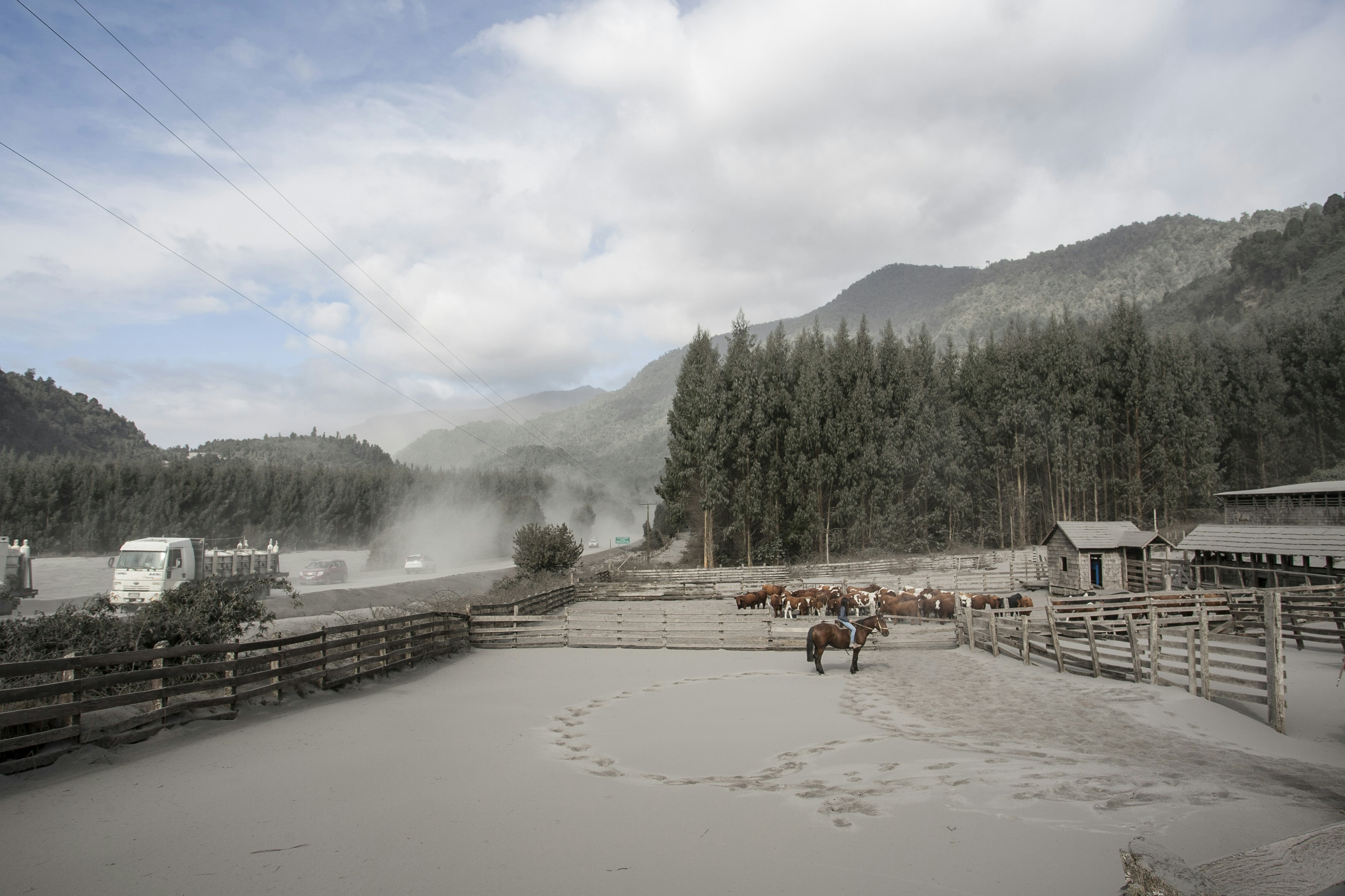 Dust plume near a rural farm yard with a herd of horses and wooden fences, set against a backdrop of mountains and a distant road. A calm, wide-shot capture of a working ranch scene.