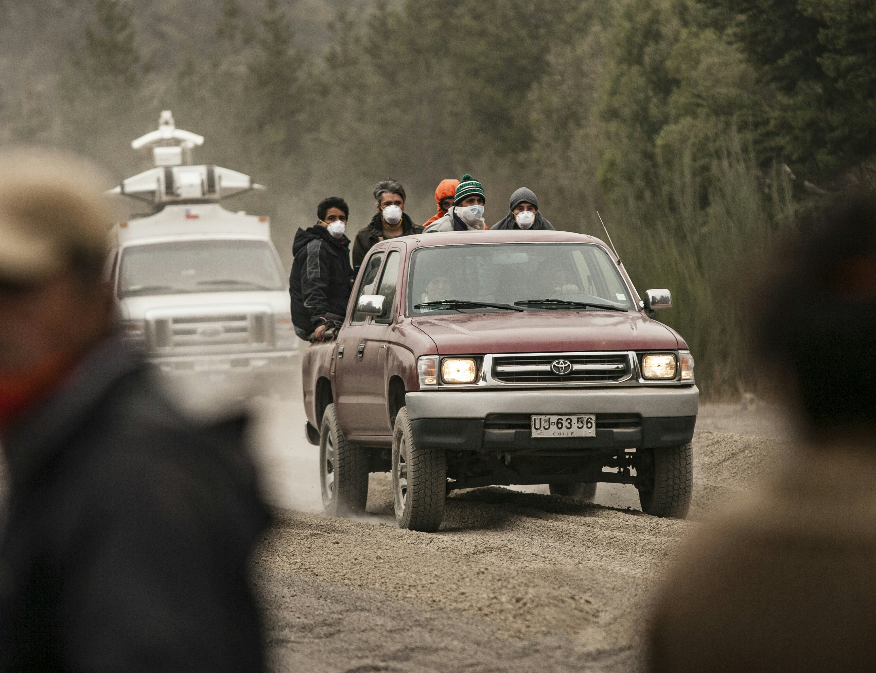 Dusty red pickup truck leads a convoy along a gravel road, with masked figures riding or walking beside and behind, while a blurred crowd frames the foreground and a forest backdrop looms; scene suggests a cautious transport or expedition.