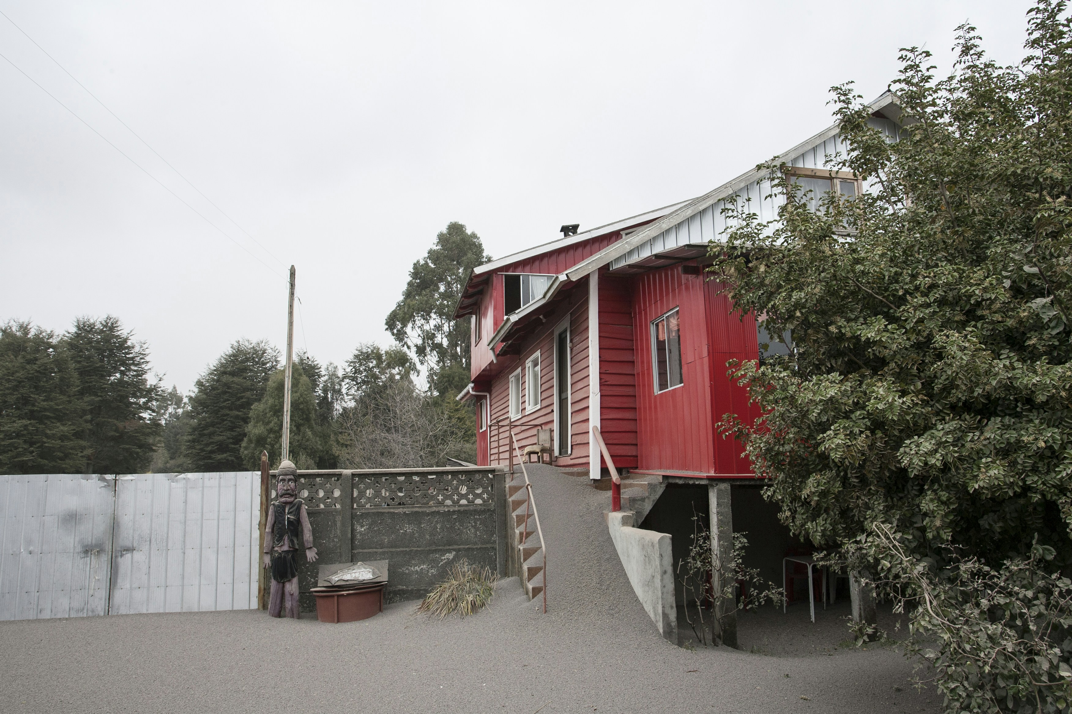 A red wooden house on stilts sits above a gravel yard, with a figure standing near a corrugated fence to the left; the scene conveys a quiet, rural everyday moment. The image appears to be a photograph of a modest dwelling with surrounding trees and a utilitarian yard.